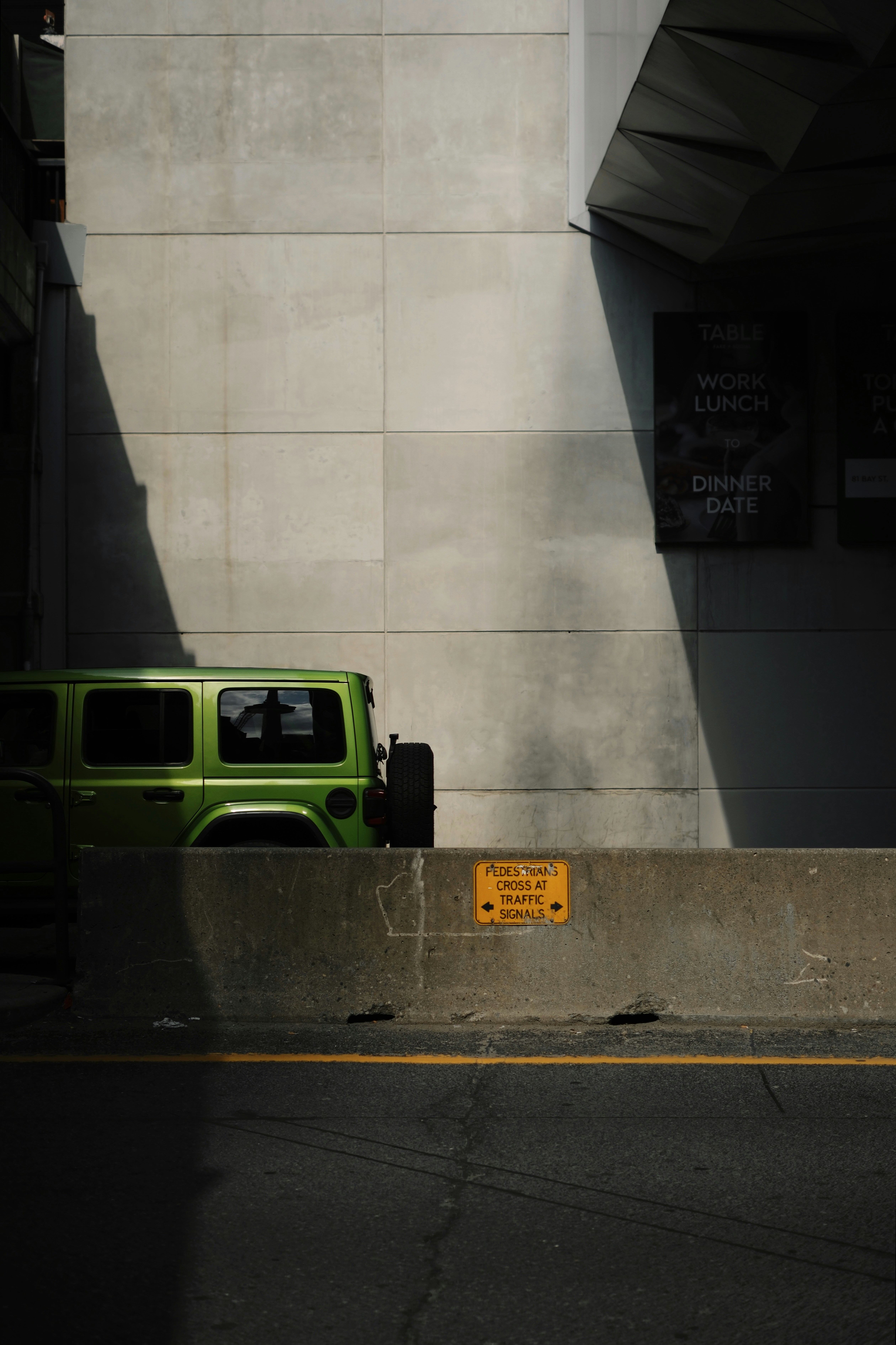 A vibrant green vehicle is parked beside a stark concrete wall, casting a long shadow. A cautionary sign is visible in the foreground.
