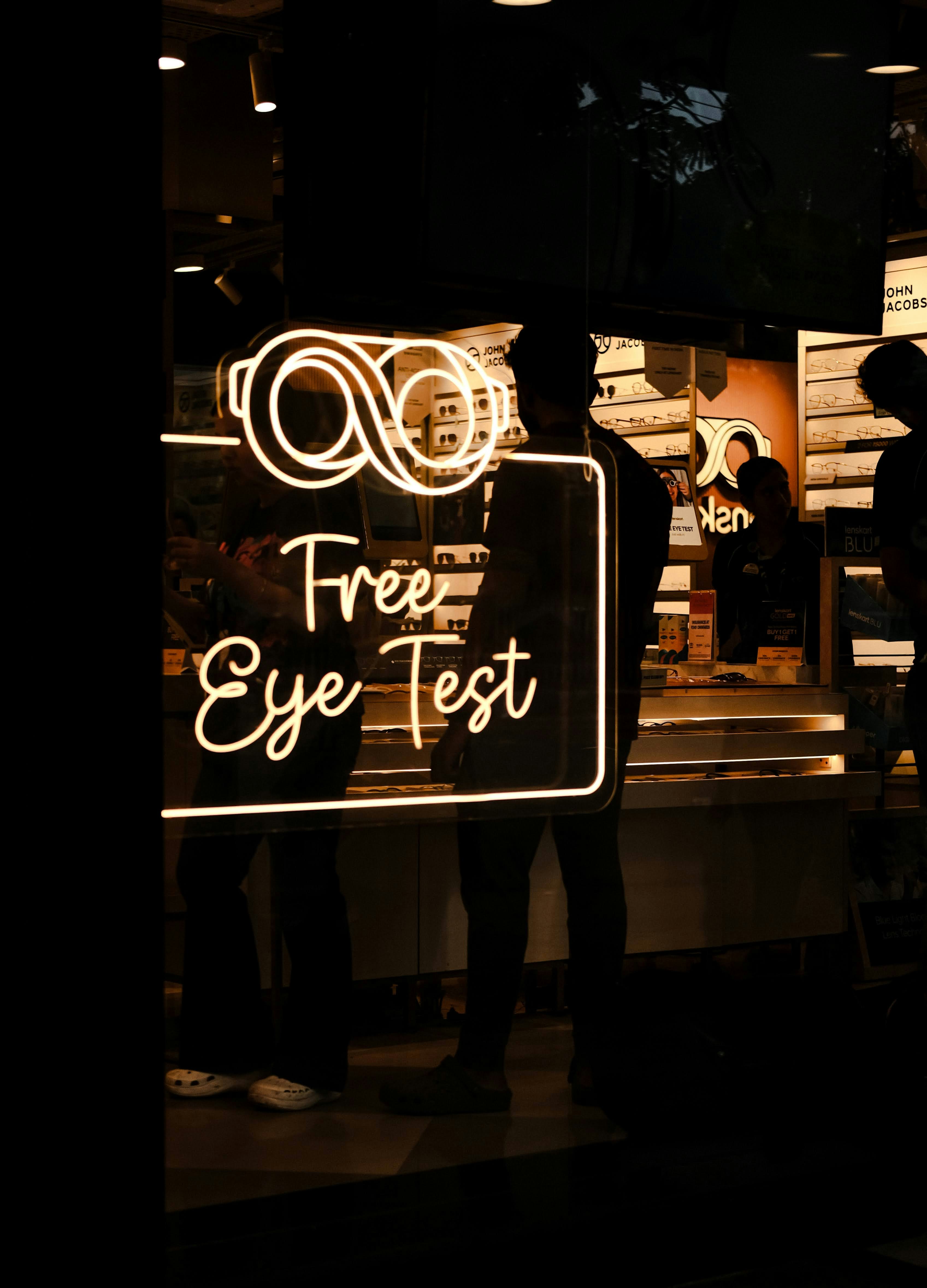 Neon sign advertising a free eye test, framed by silhouettes of people inside an optical store. The glow of the sign contrasts with the dim interior.