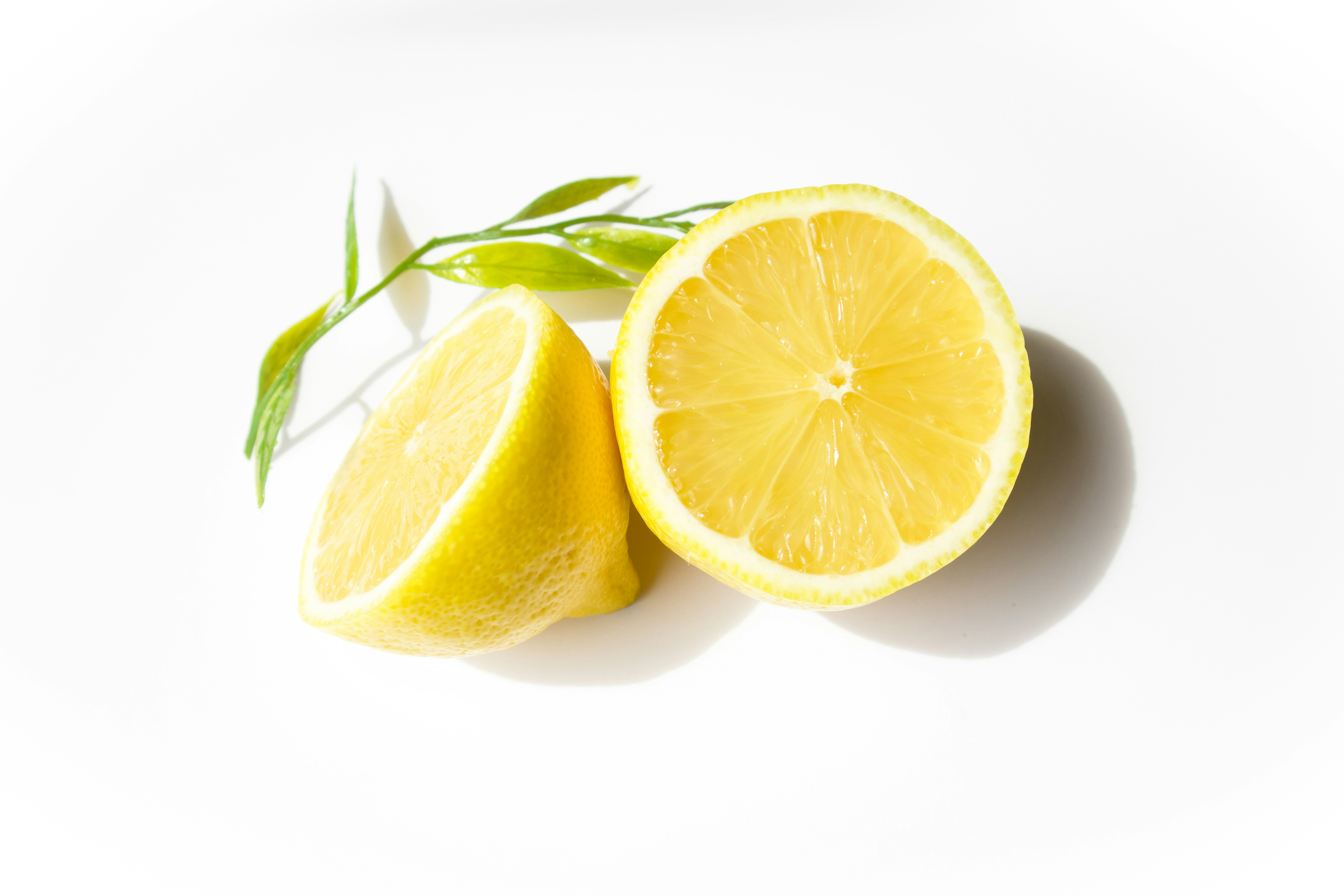 A pair of fresh lemon halves rest on a pristine white surface, their vibrant yellow flesh glistening in the sunlight. The composition is accented by a delicate sprig of green leaves, creating a visually refreshing and clean aesthetic. The strong, natural light casts soft shadows, highlighting the juicy texture and inviting viewers to imagine the citrus aroma. This image is perfect for illustrating concepts of freshness, summer, healthy eating, or natural ingredients, making it ideal for food blogs, advertisements, or wellness content.