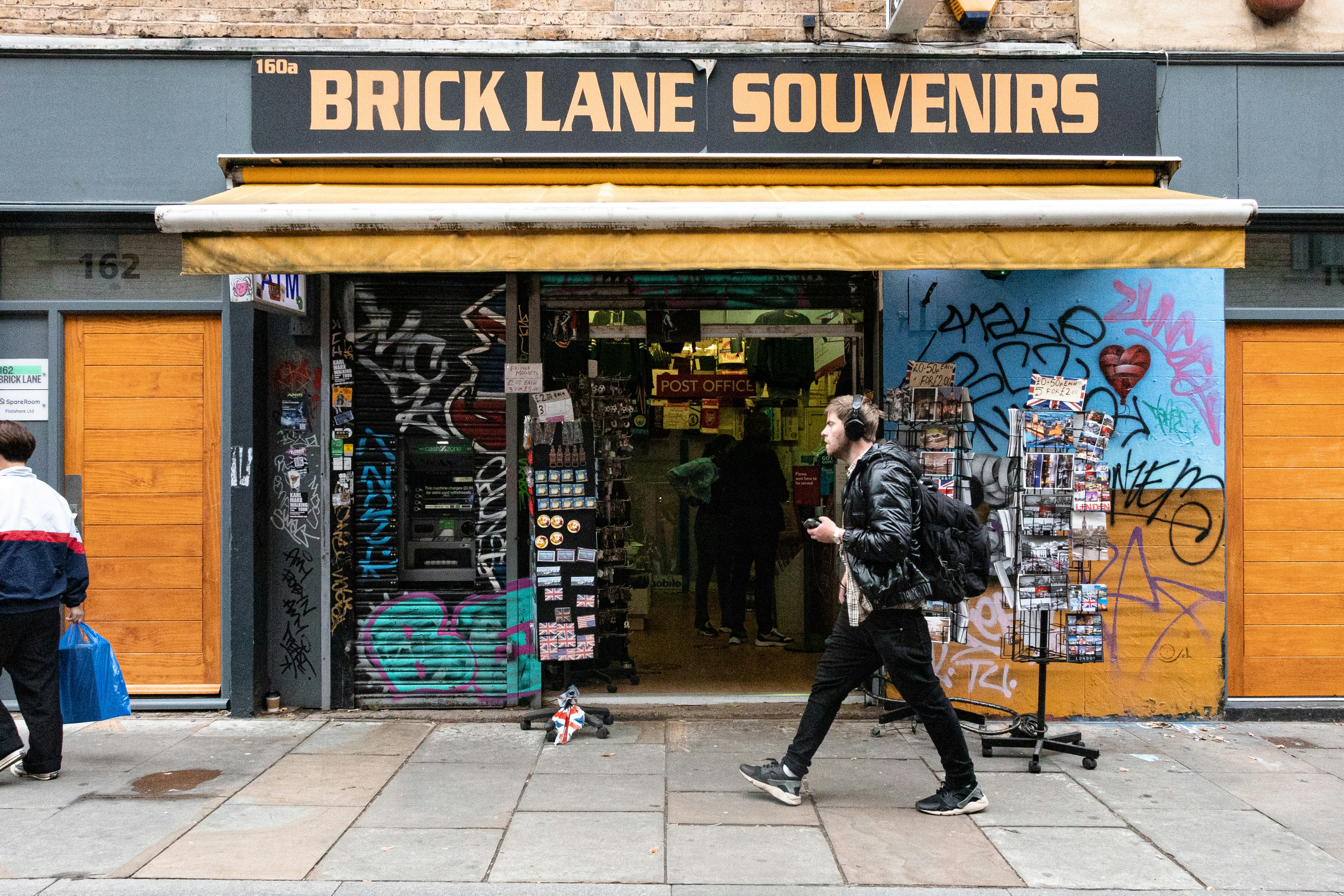 Colorful graffiti adorns the exterior of a souvenir shop, with a passerby engaged in their surroundings. The vibrant urban scene reflects the eclectic spirit of Brick Lane.