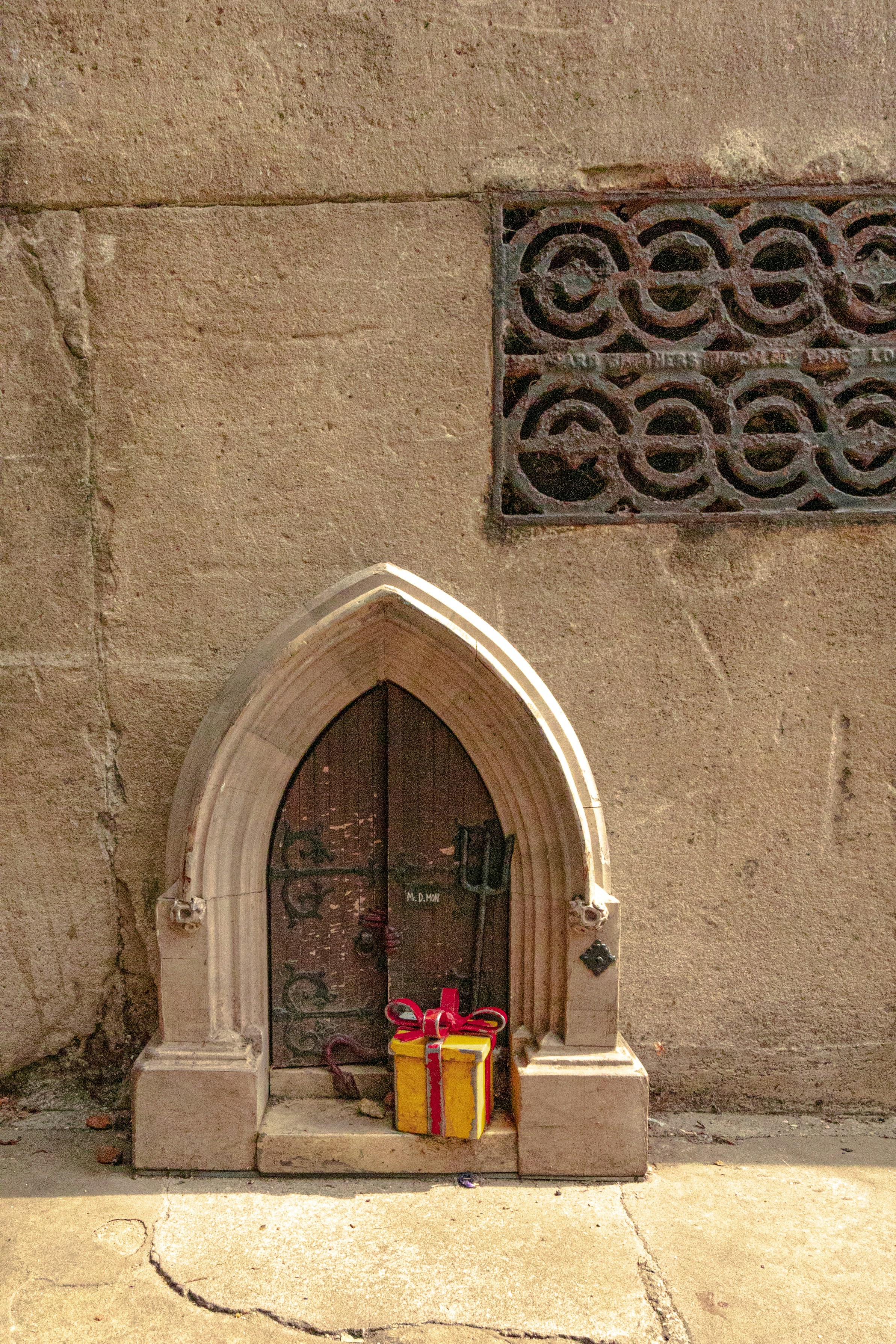 A small, intricately carved door set into a stone wall, accompanied by a brightly wrapped gift at its base.