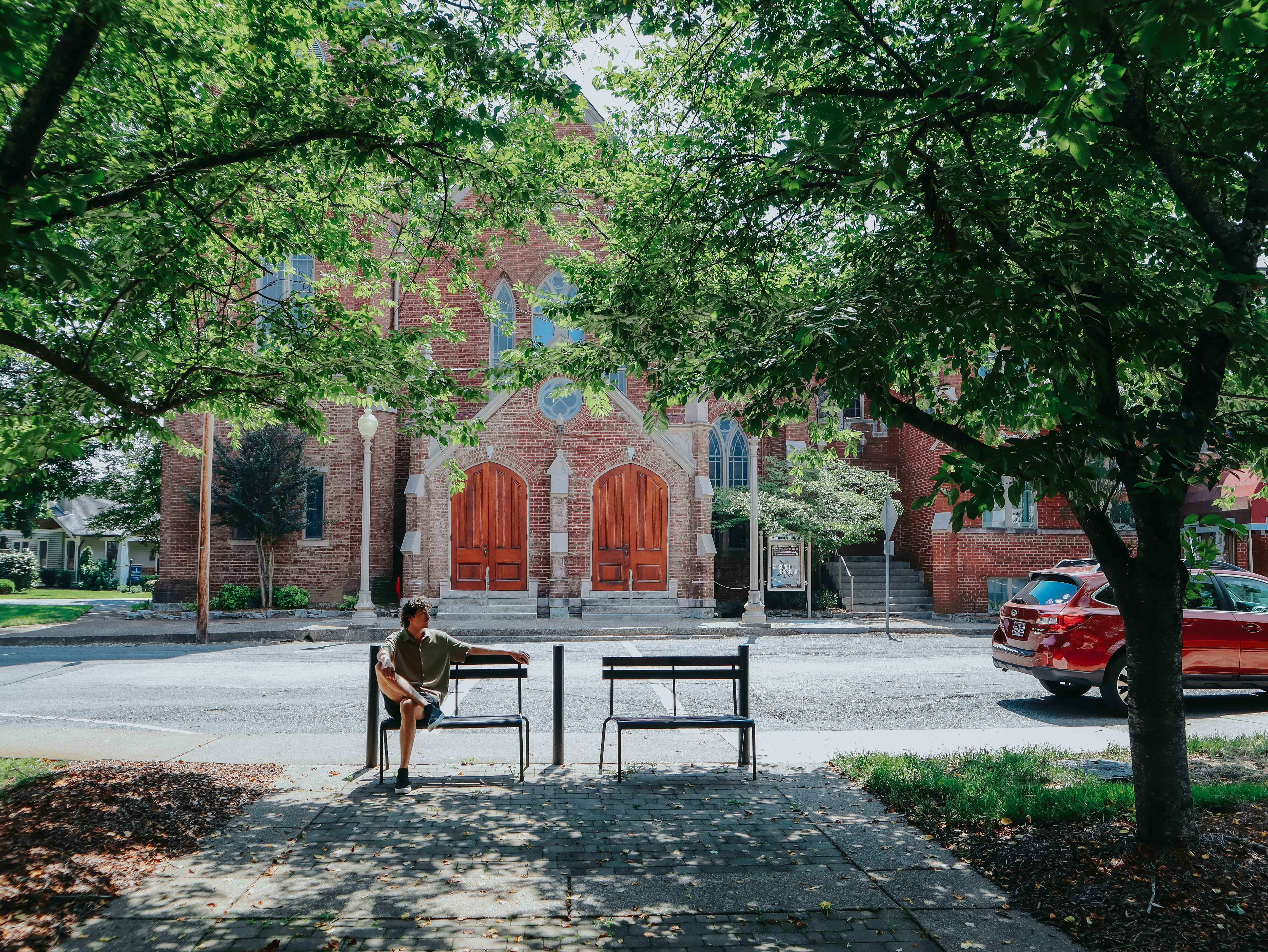 Individual seated on a bench under a canopy of trees, with a church backdrop and a parked car in a quiet street scene.