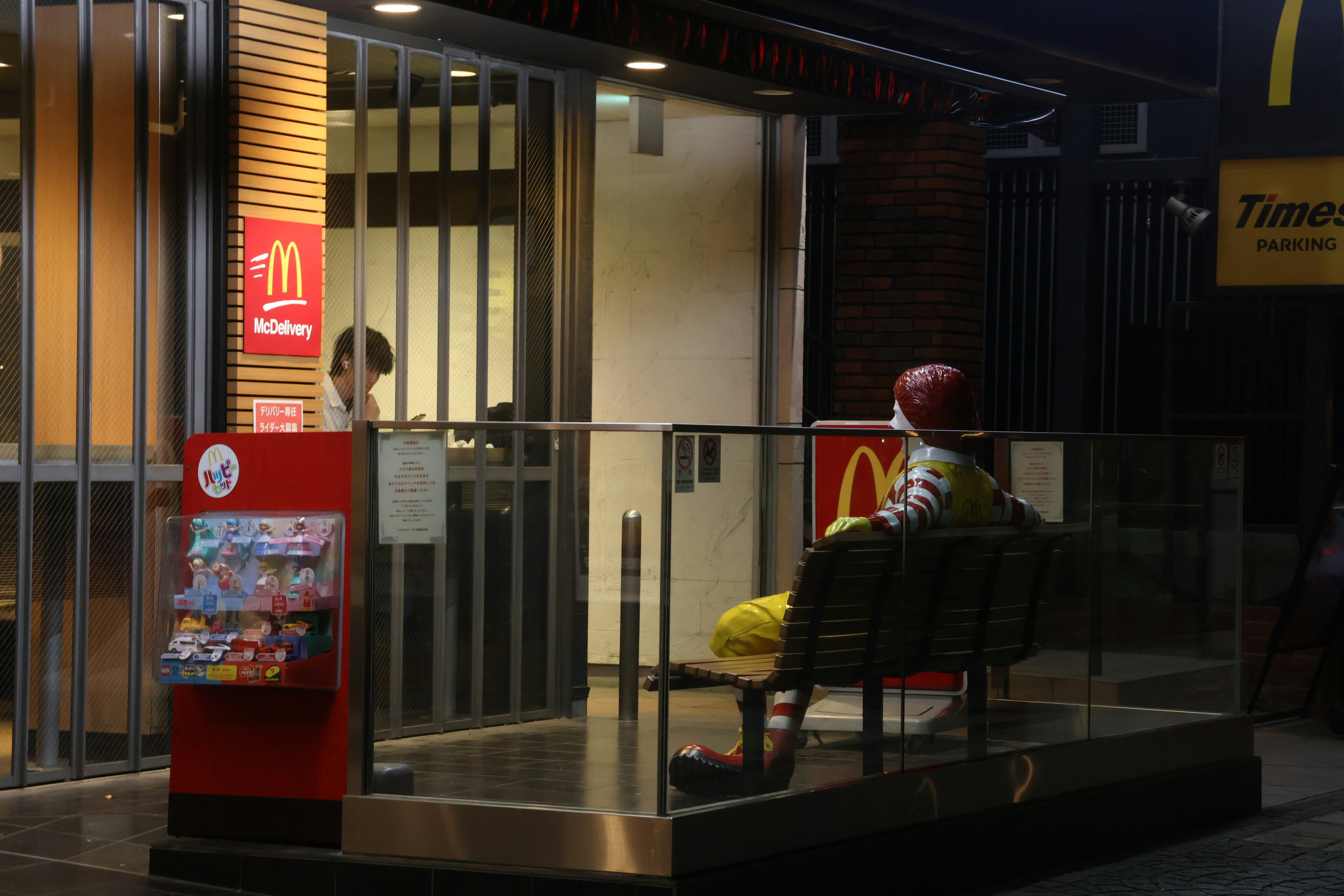 A whimsical scene featuring a Ronald McDonald statue seated on a bench outside a McDonald's restaurant, illuminated by the warm glow of the establishment's lights.