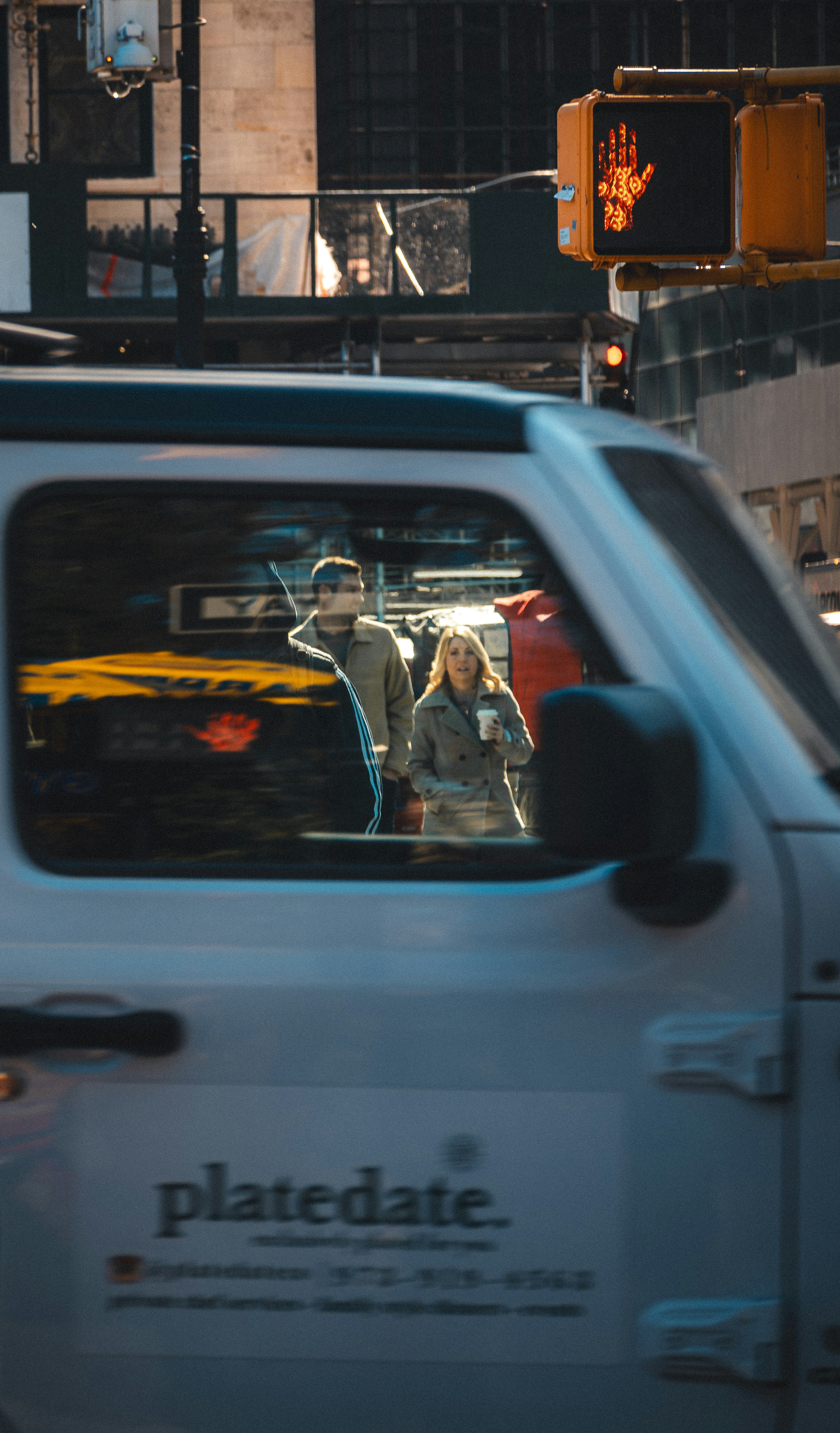 A reflective view of pedestrians waiting at a crosswalk, framed by a passing vehicle and a traffic signal displaying a hand symbol.