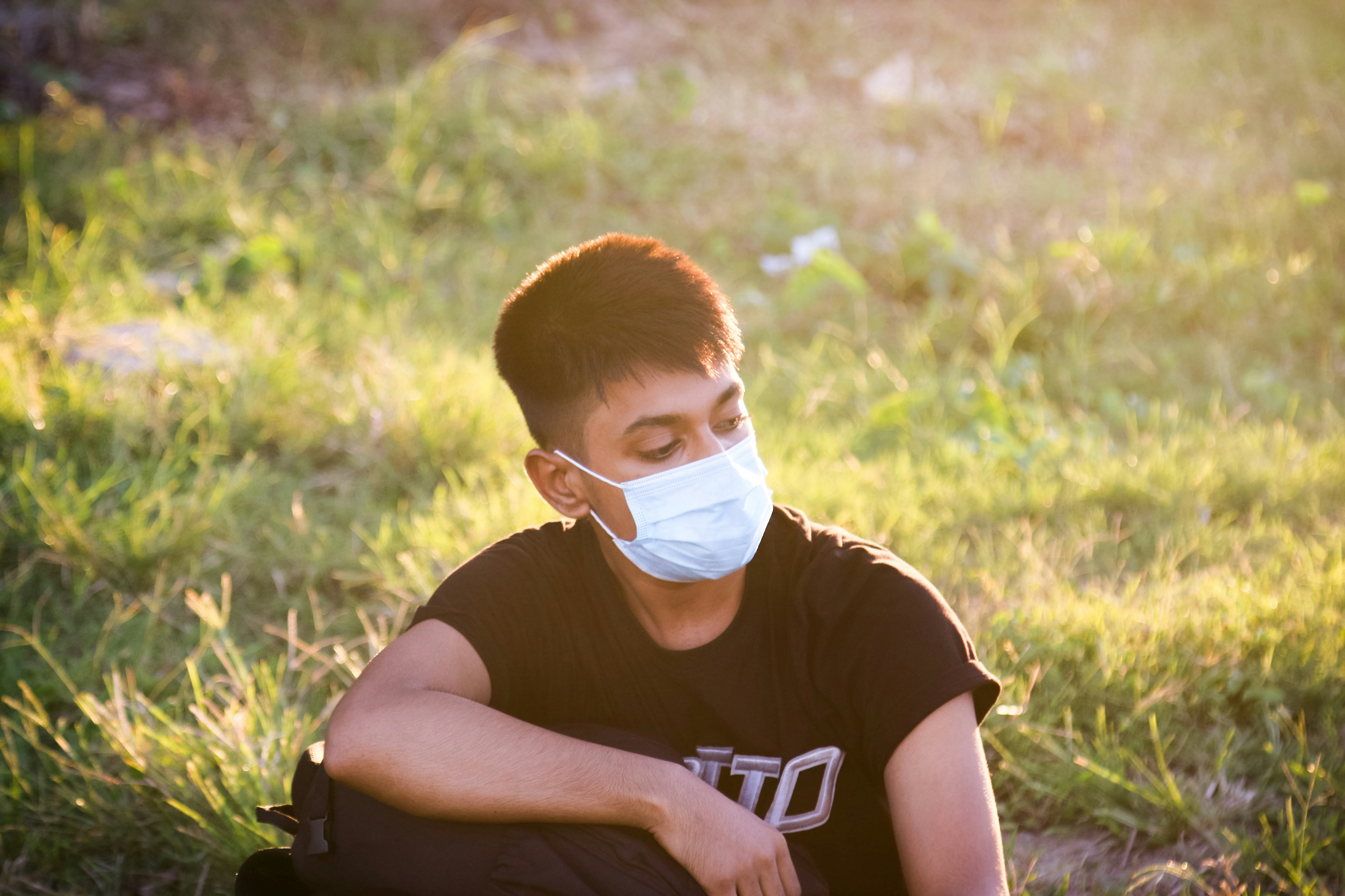 A young man wearing a face mask sits thoughtfully on a grassy field, bathed in soft natural sunlight. This image reflects the everyday moments and lifestyle shifts during the pandemic, portraying a blend of solitude, safety, and calmness in the outdoors. | A young man wears a mask outdoors.