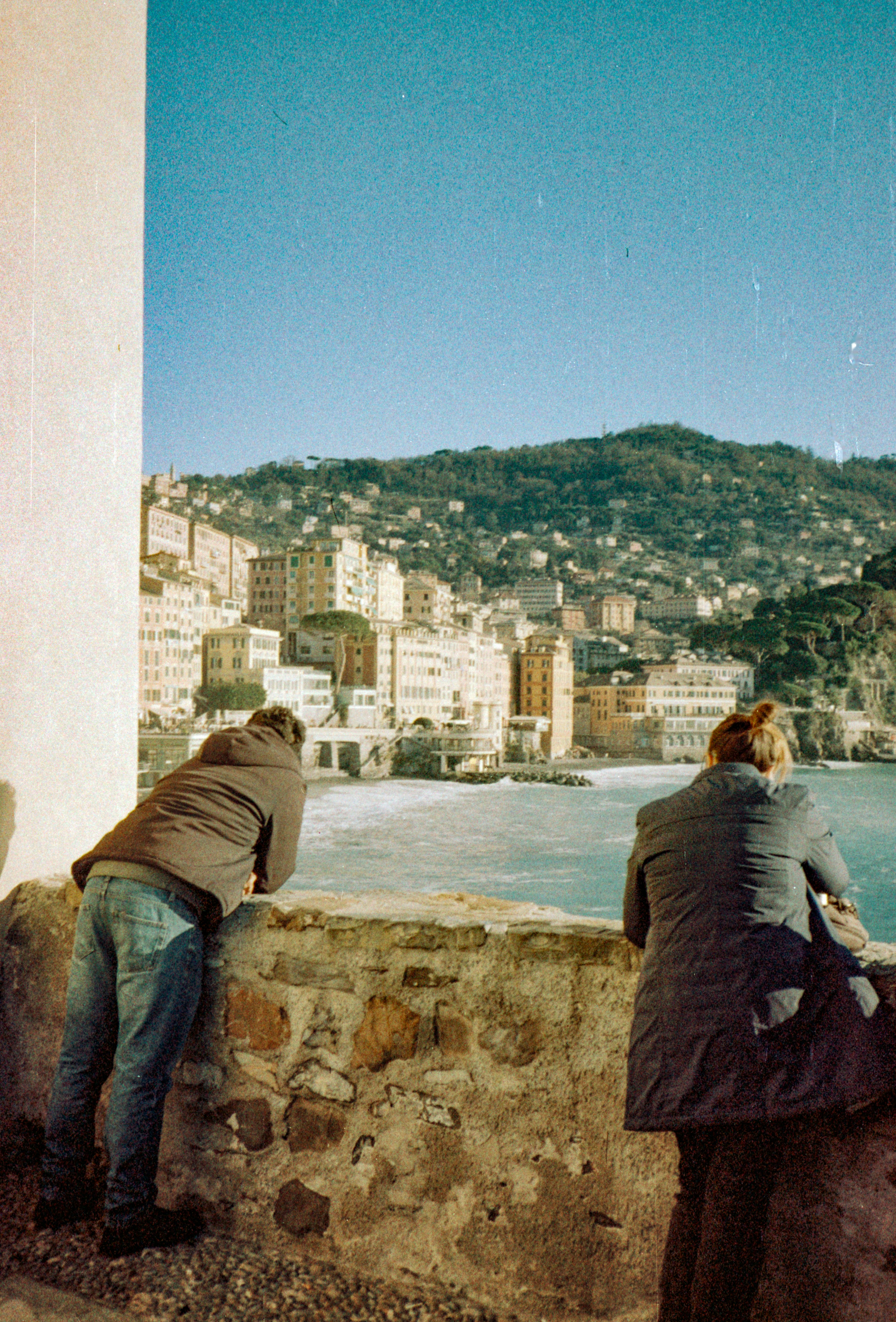 Two individuals leaning over a stone railing, gazing at the tranquil coastal scenery with colorful buildings nestled against a hillside.
