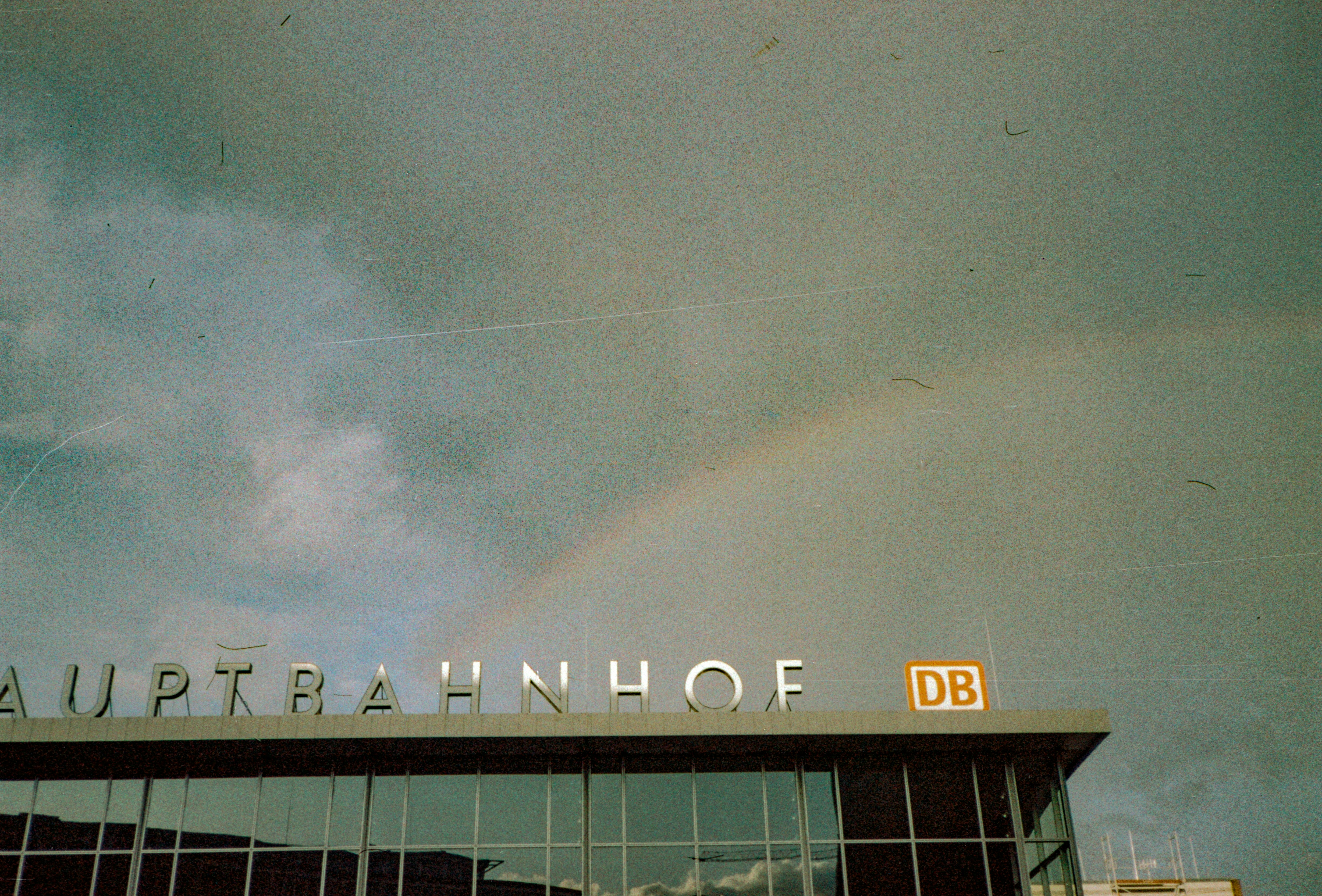 Rainbow arches over the Hauptbahnhof station, reflecting the interplay of light and architecture. The scene captures a moment of tranquility amidst urban life.