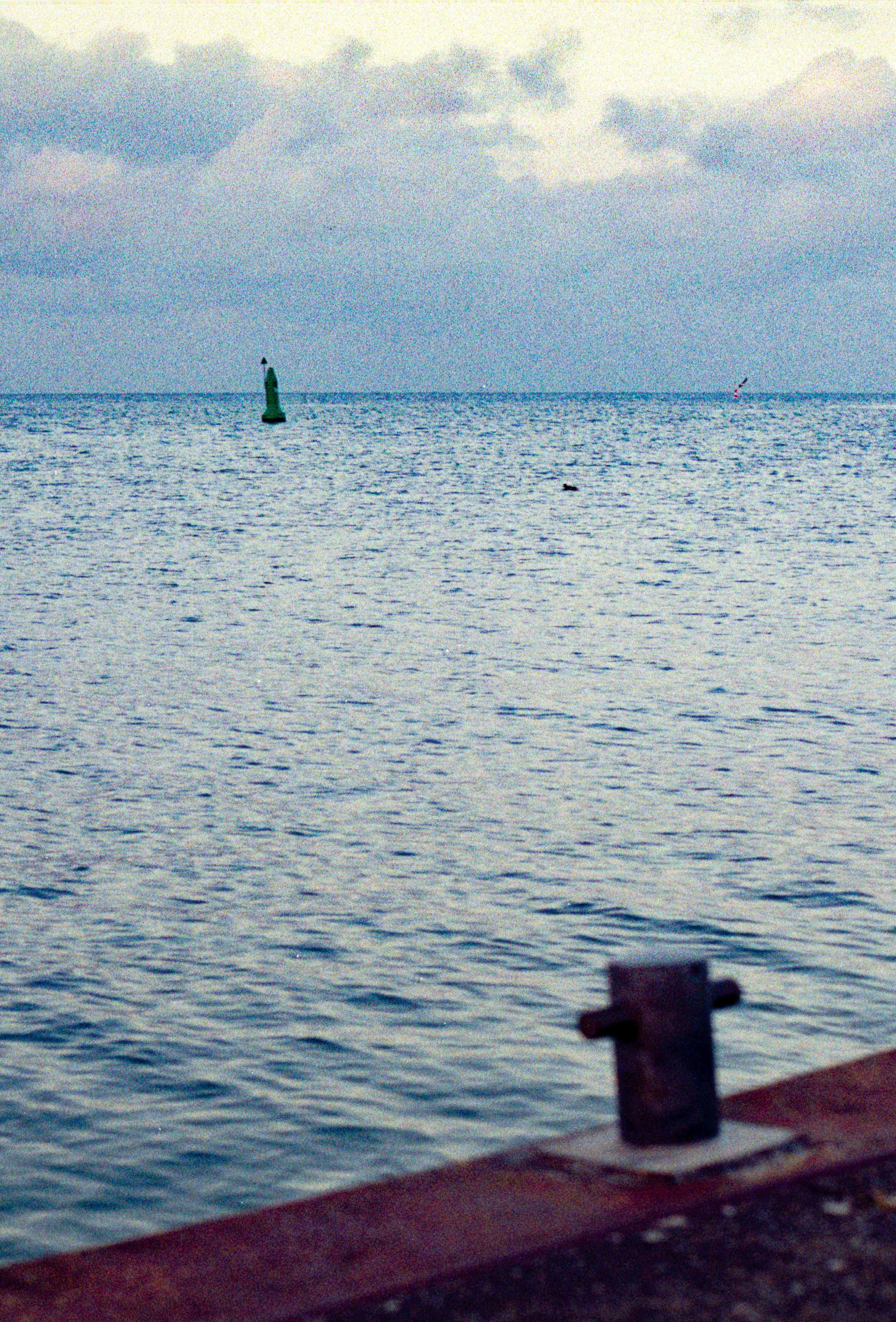 Rusty mooring post in the foreground with a distant green buoy on calm water under a cloudy sky.