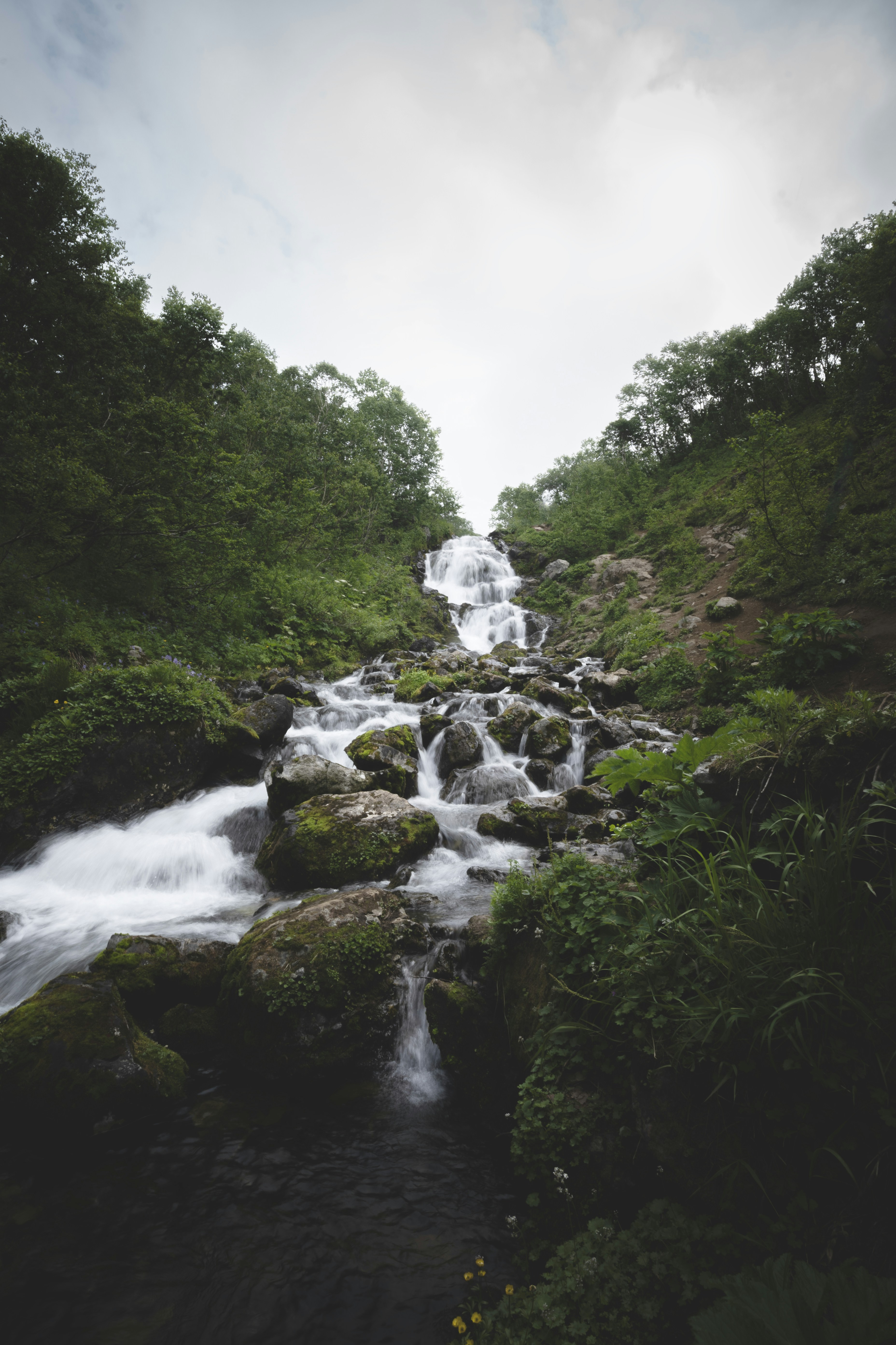 A tranquil waterfall cascading over moss-covered rocks, surrounded by lush greenery under a cloudy sky.