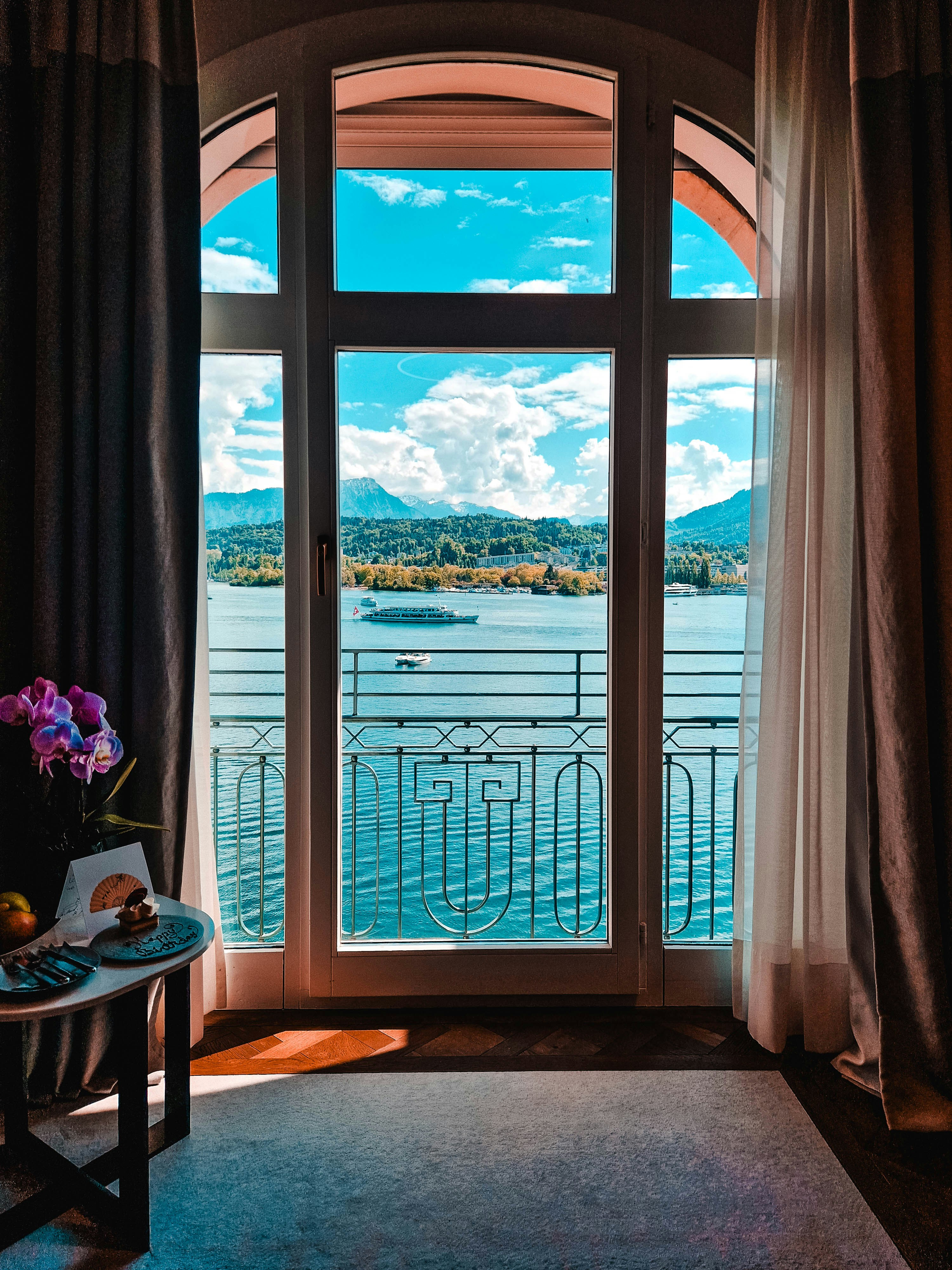 View of a tranquil lake through an elegant window, framed by soft curtains and a decorative railing. A boat glides across the water under a bright blue sky.