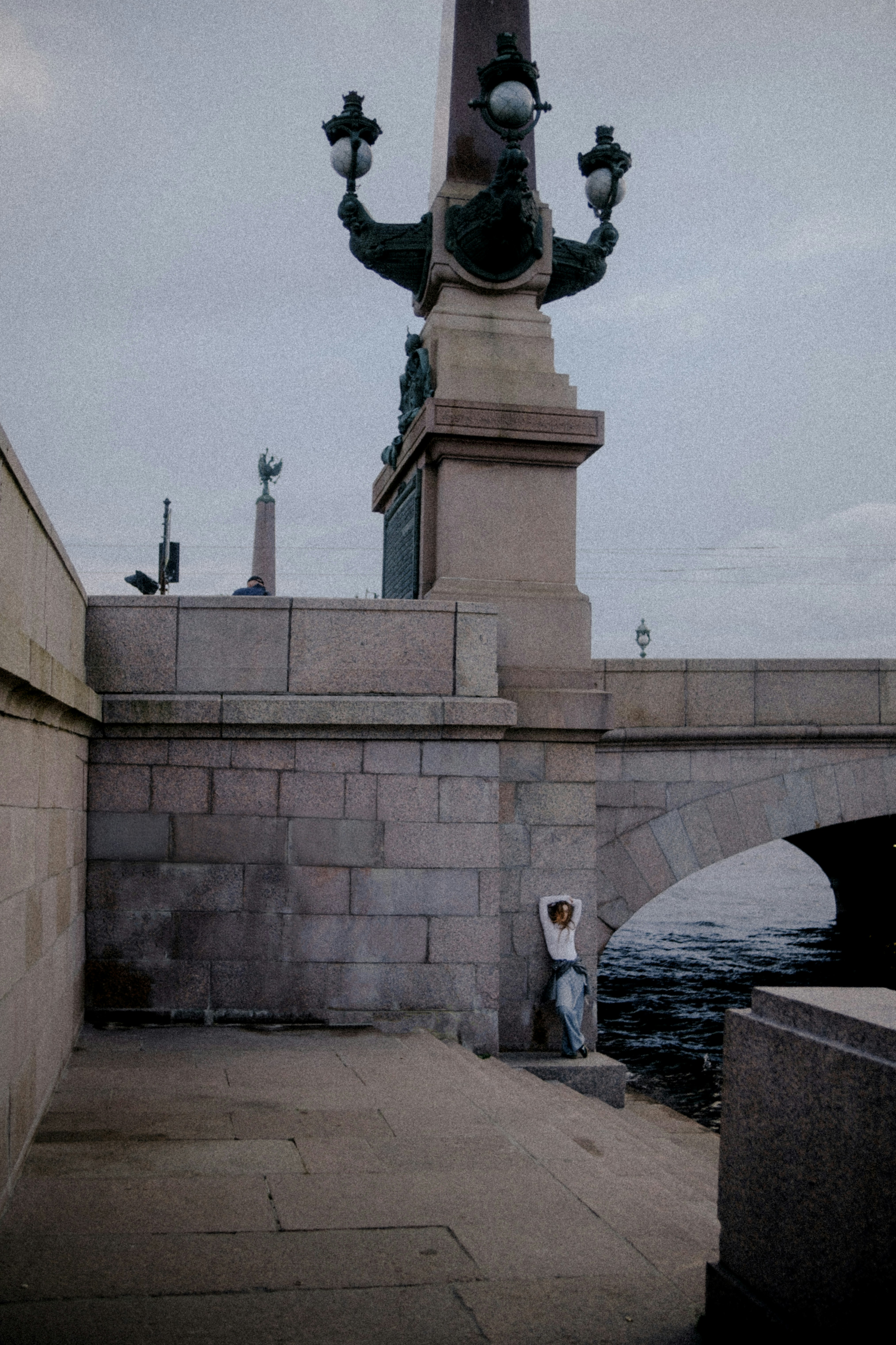 Lone Figure by Stone Bridge Lamps: Echoes Along the Riverfront