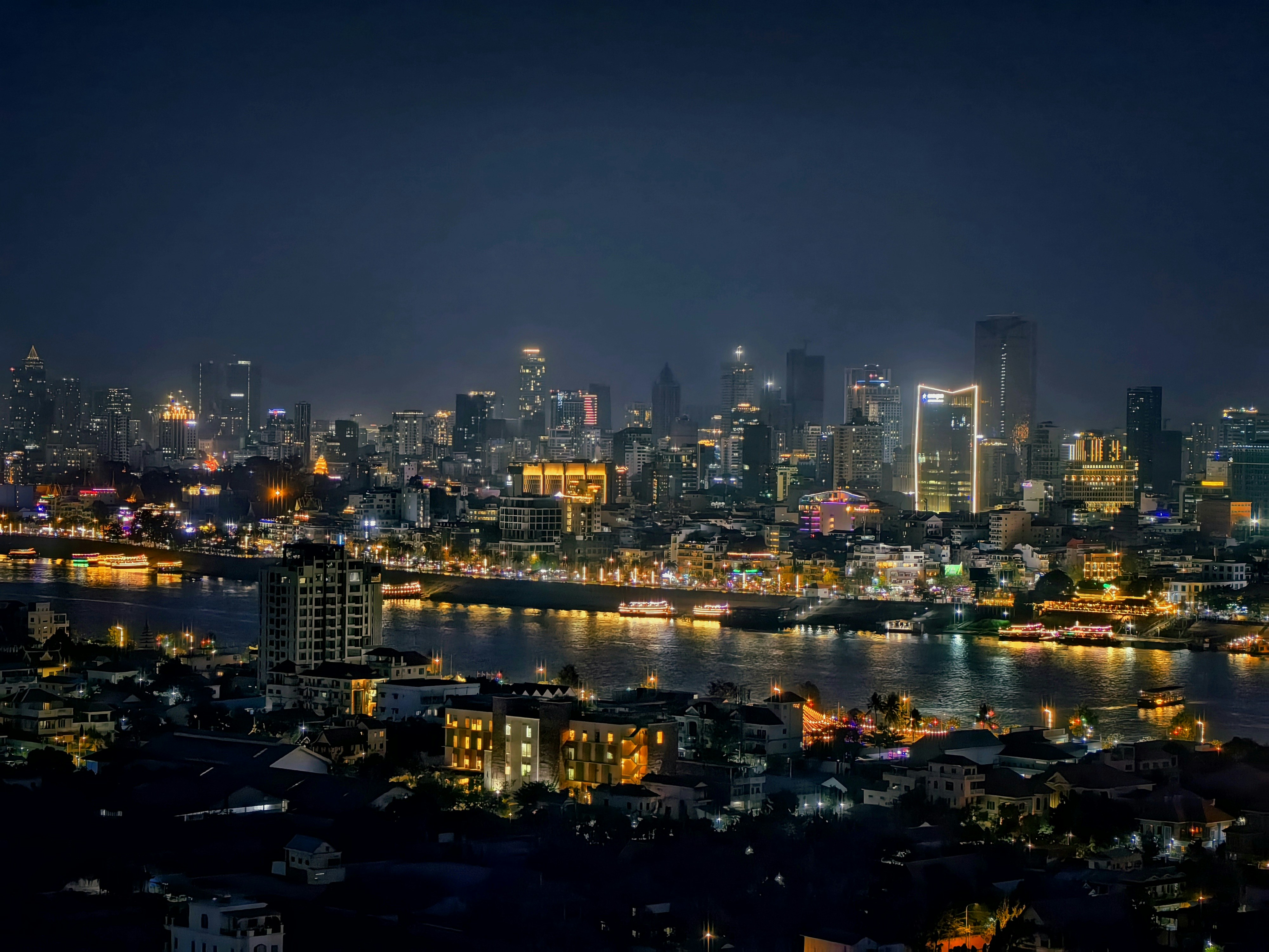 Vibrant city skyline illuminated by countless lights, reflecting on the calm river below at night.