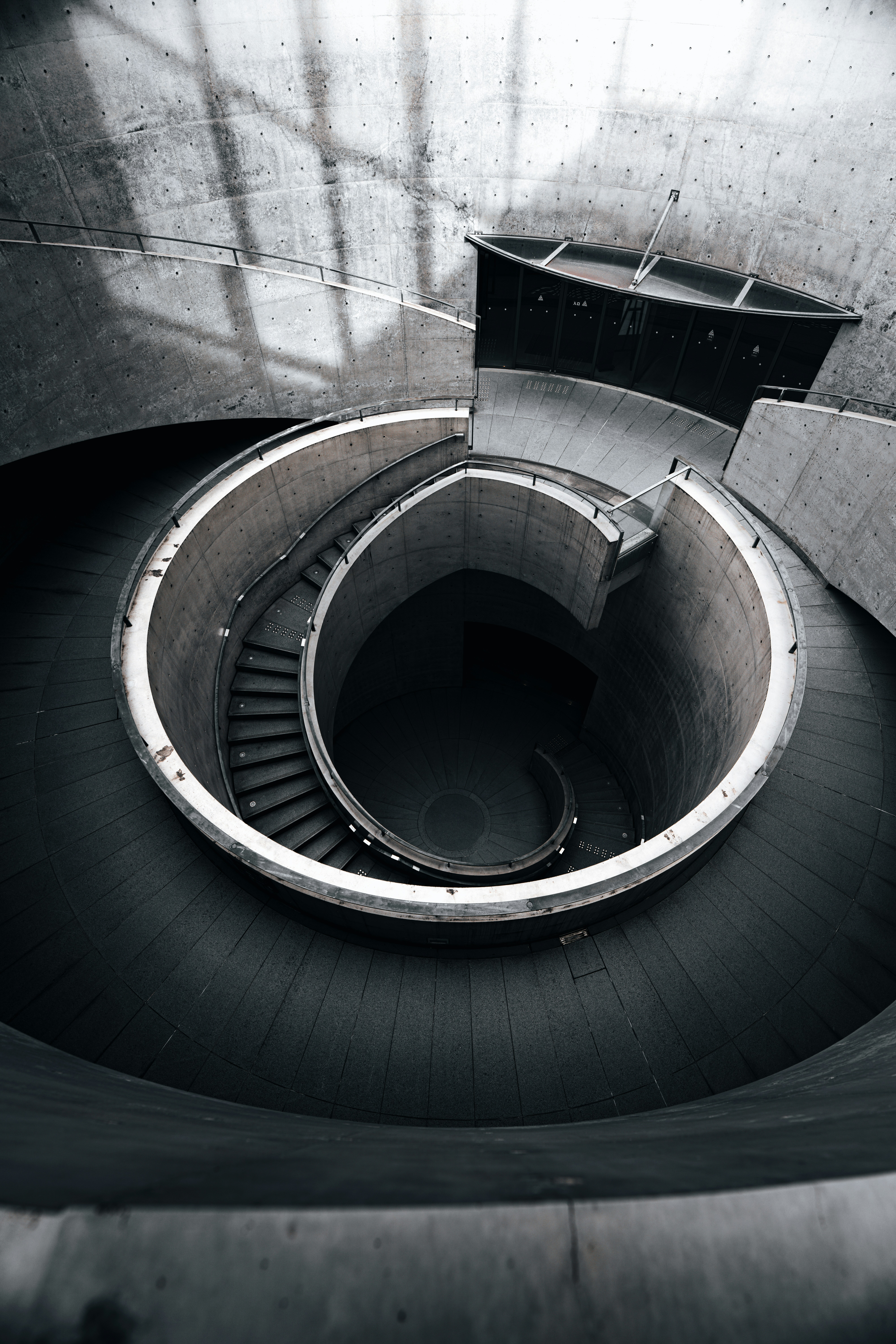 A spiraling staircase within a minimalist concrete structure, showcasing modern architectural design. The circular form draws the eye towards the center.