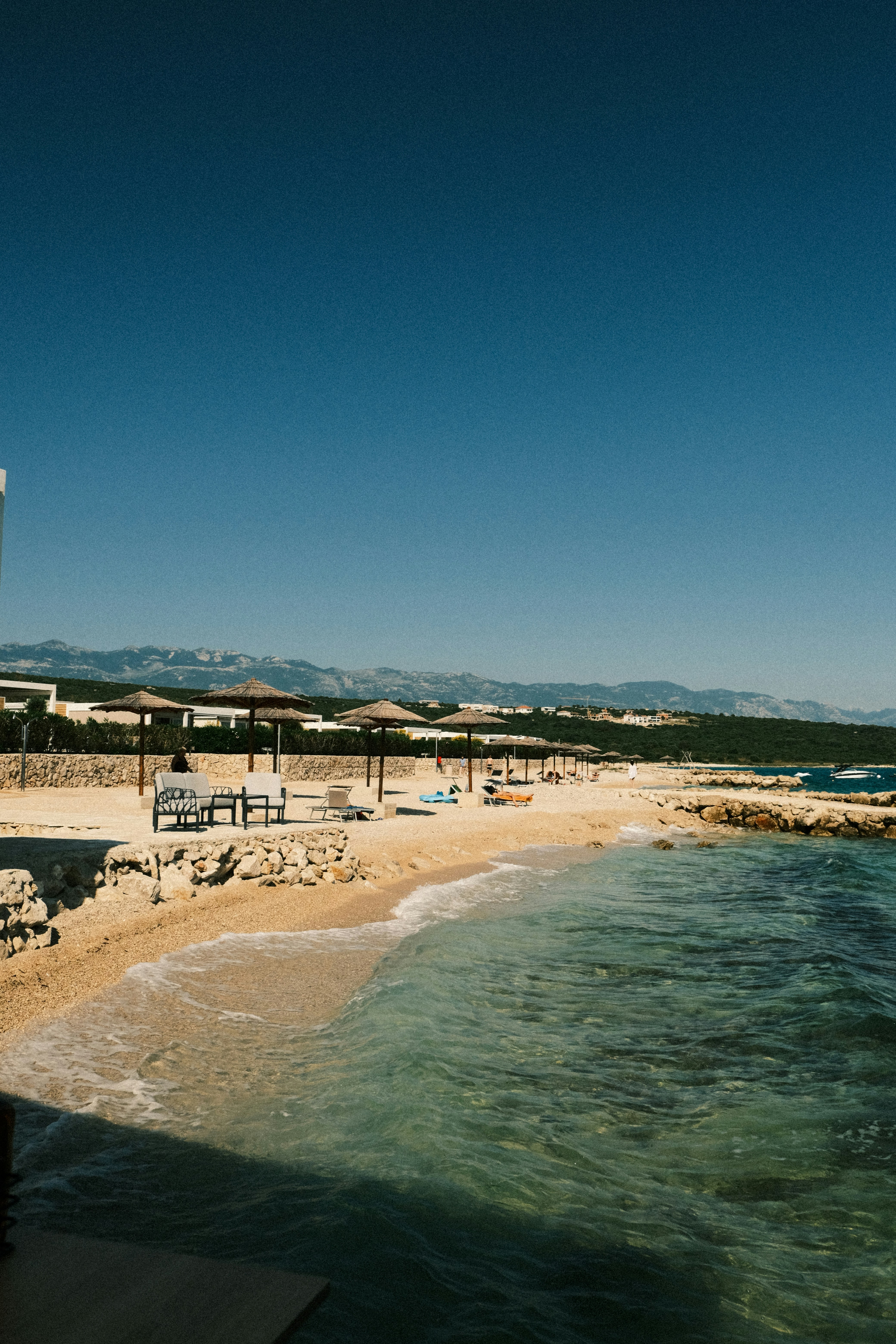 Tranquil beach scene featuring sandy shores, shaded loungers, and clear blue waters under a bright sky.