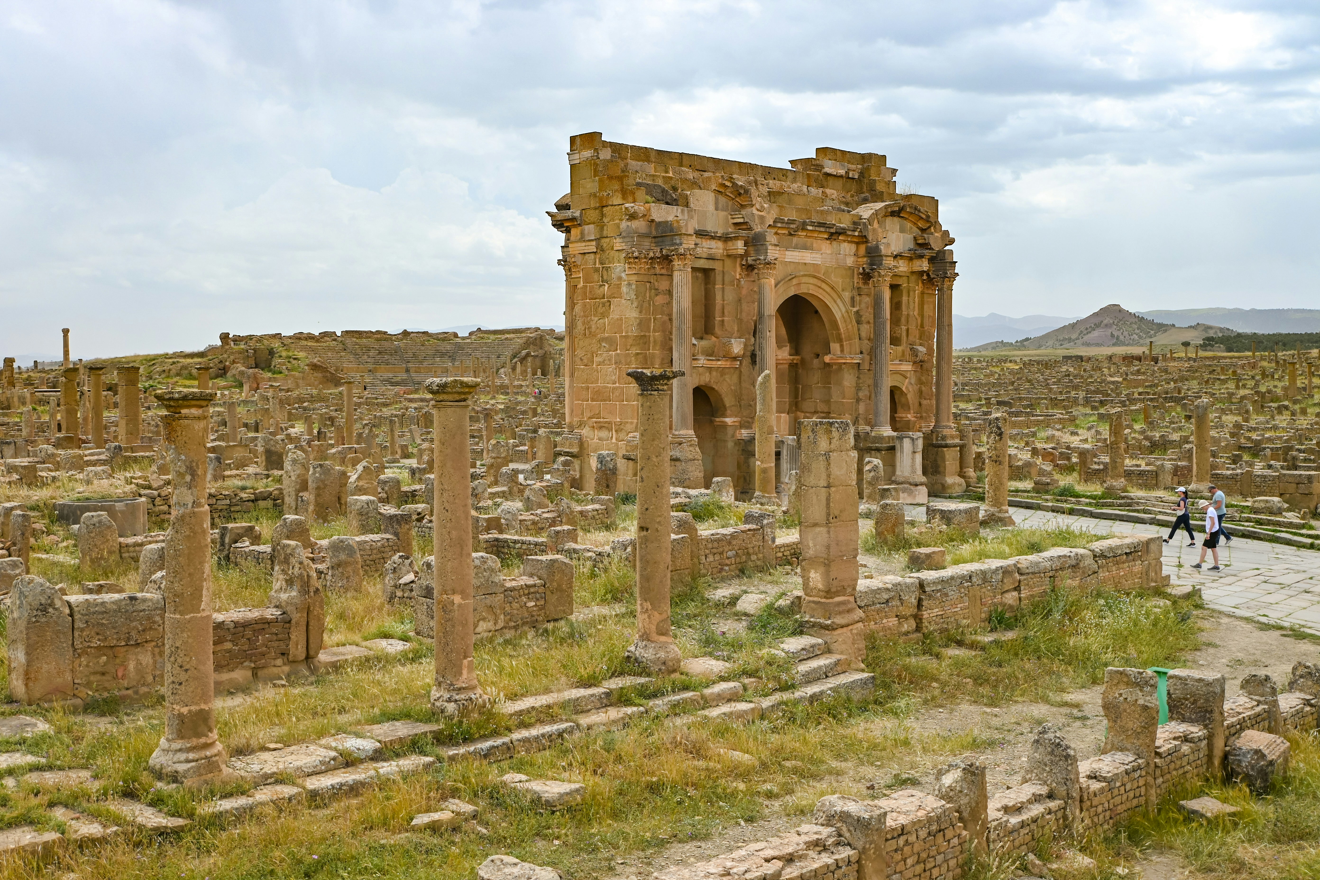 Ancient Roman ruins of Timgad with a prominent archway and scattered columns under a cloudy sky.