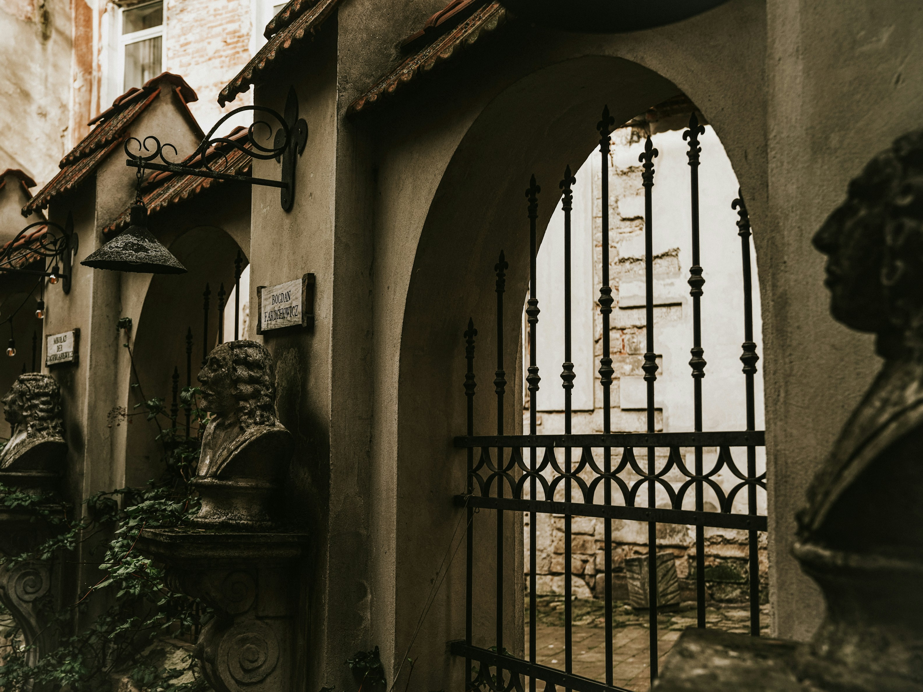 Stone busts line a weathered wall, framed by wrought iron gates and lush greenery, suggesting a forgotten garden steeped in history.