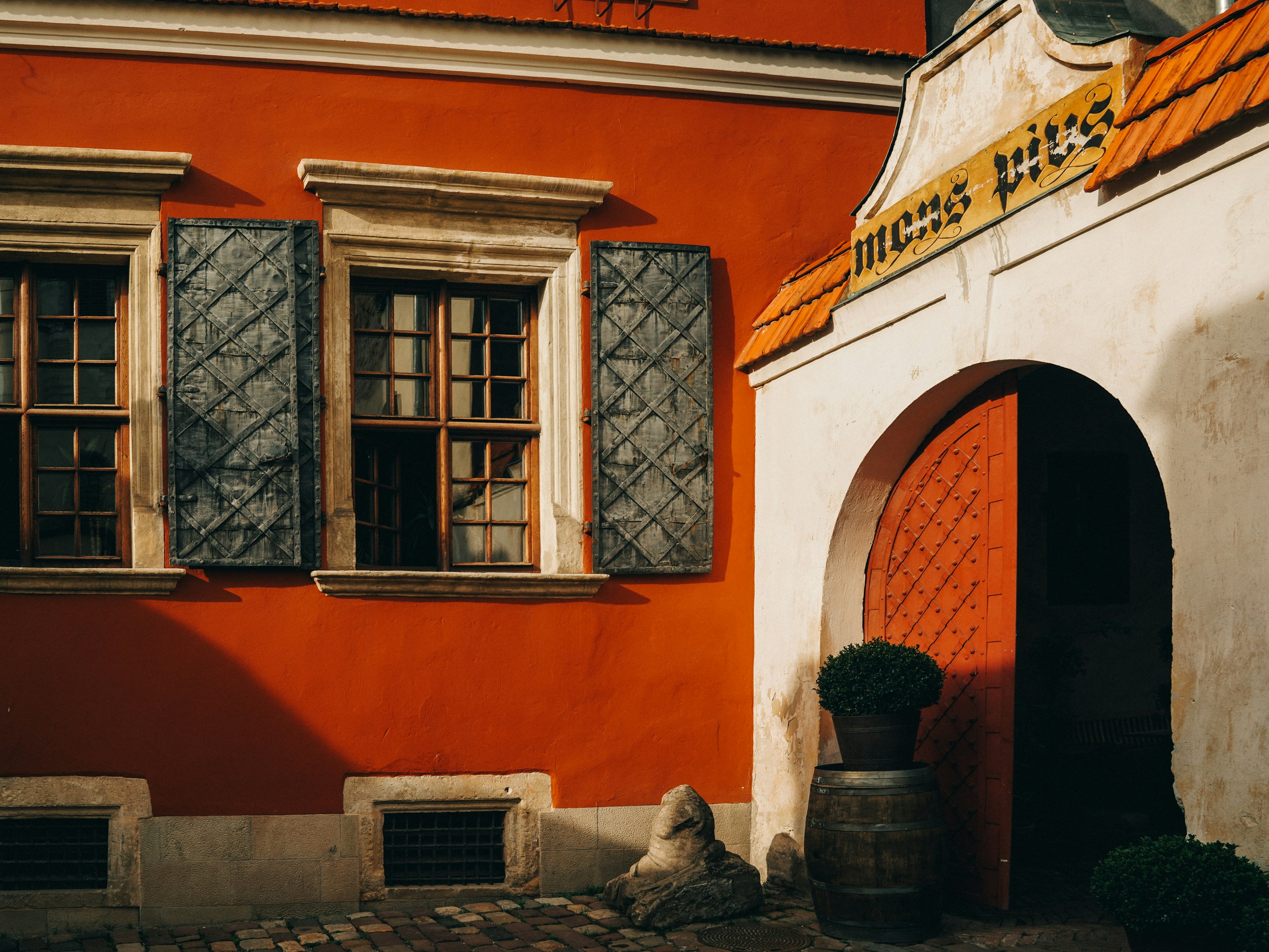 Charming orange wall adorned with wooden shutters and a welcoming archway, revealing a glimpse of a rustic entrance. A stone figure rests nearby, adding character to the scene.