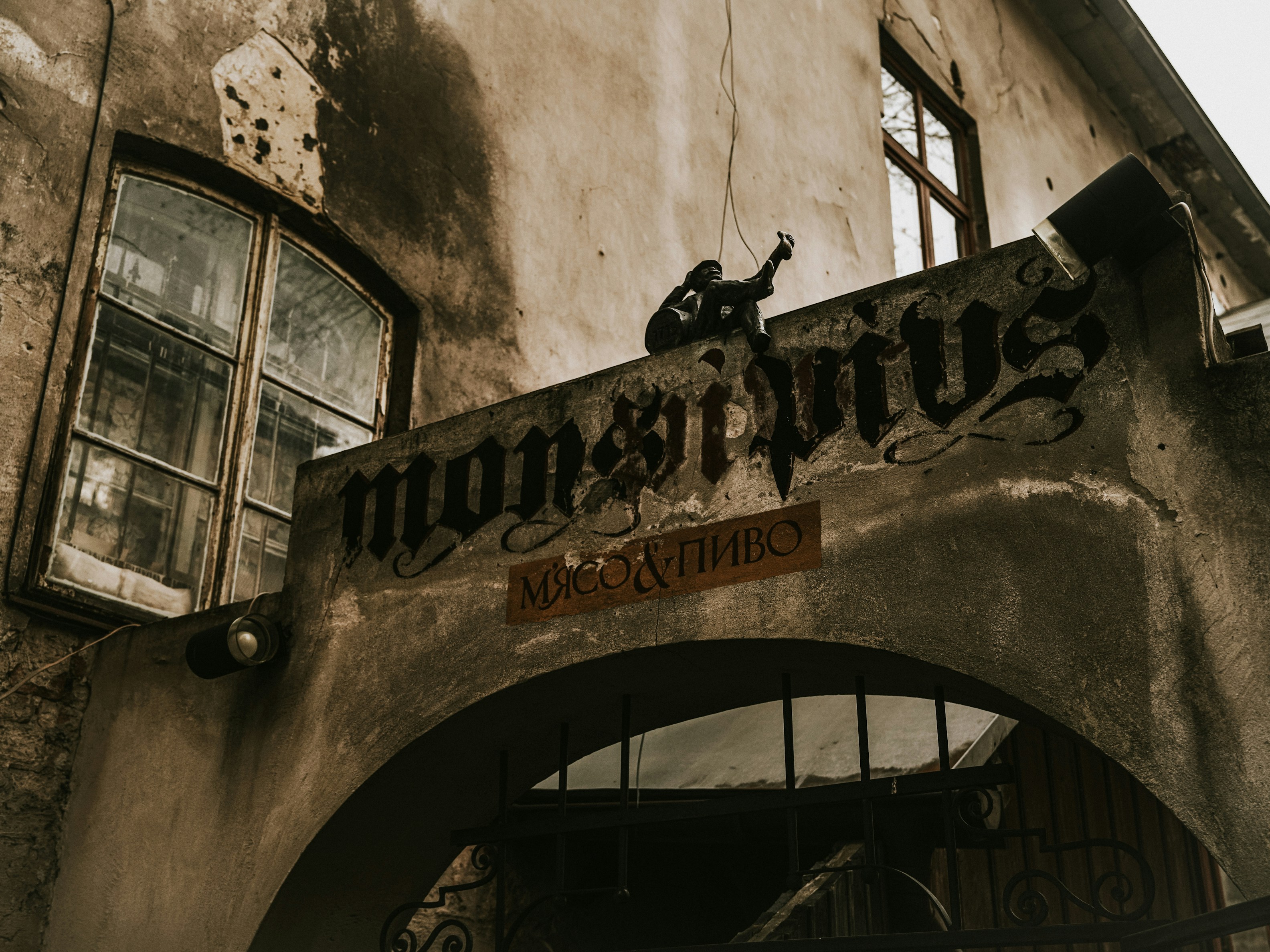 Weathered entrance to a building featuring ornate lettering and a small figure atop the archway. The scene conveys a sense of history and intrigue.