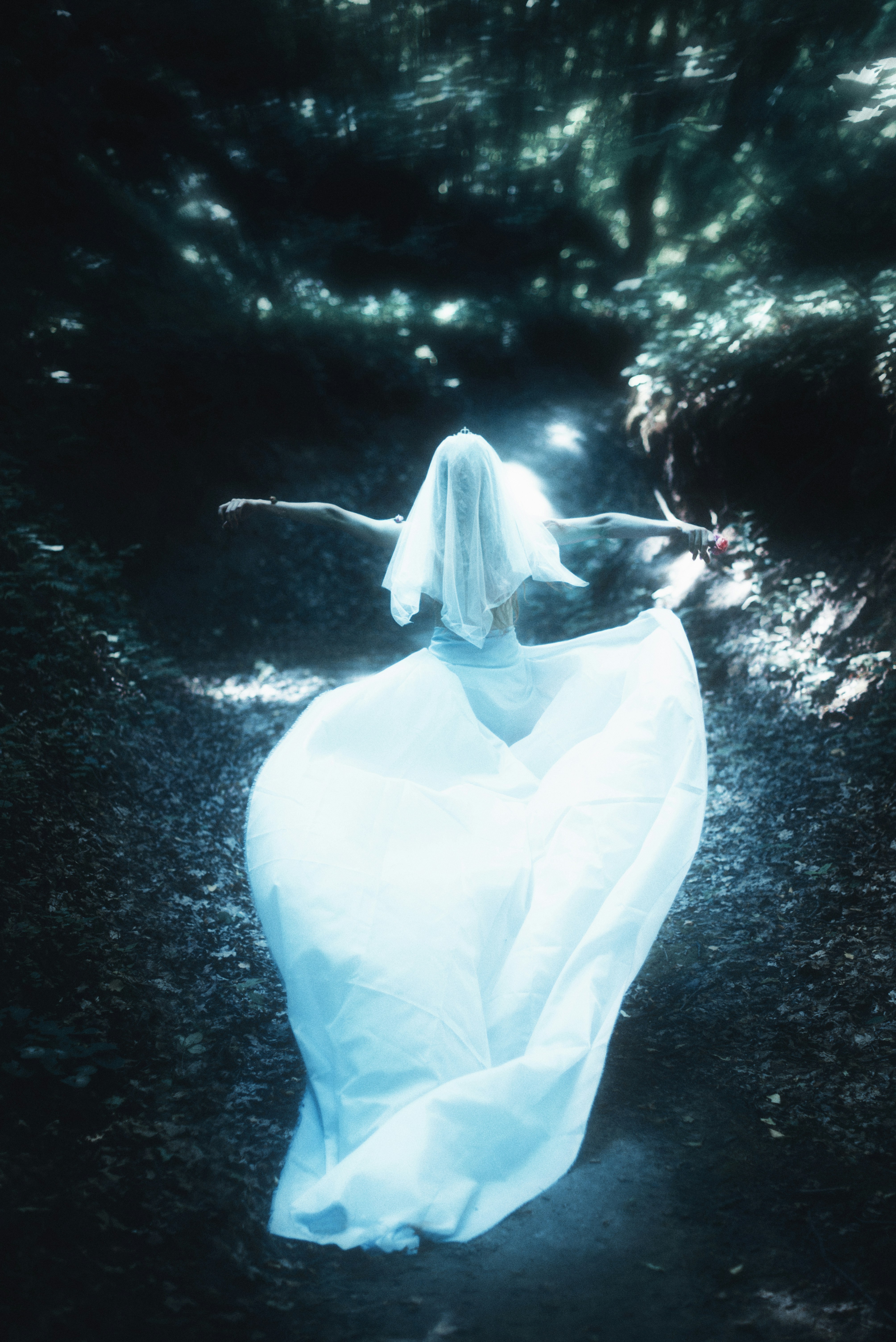 Bride in flowing white gown and veil walking through a shadowy forest, holding a bouquet. Ethereal light filters through the trees, enhancing the mystical atmosphere.