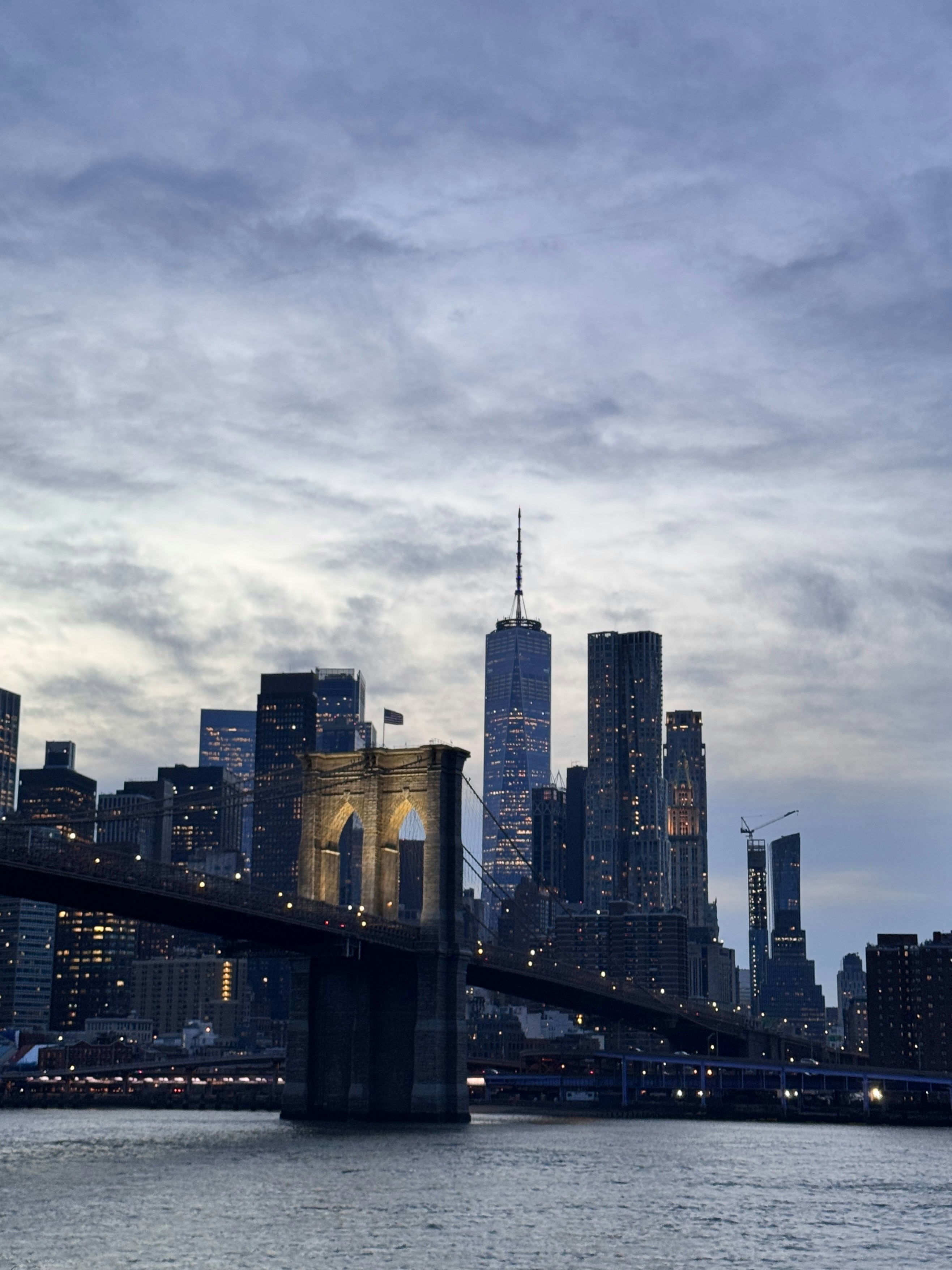Brooklyn Bridge illuminated against a backdrop of Manhattan skyscrapers during twilight. The scene captures the transition from day to night with vibrant city lights reflecting in the water.