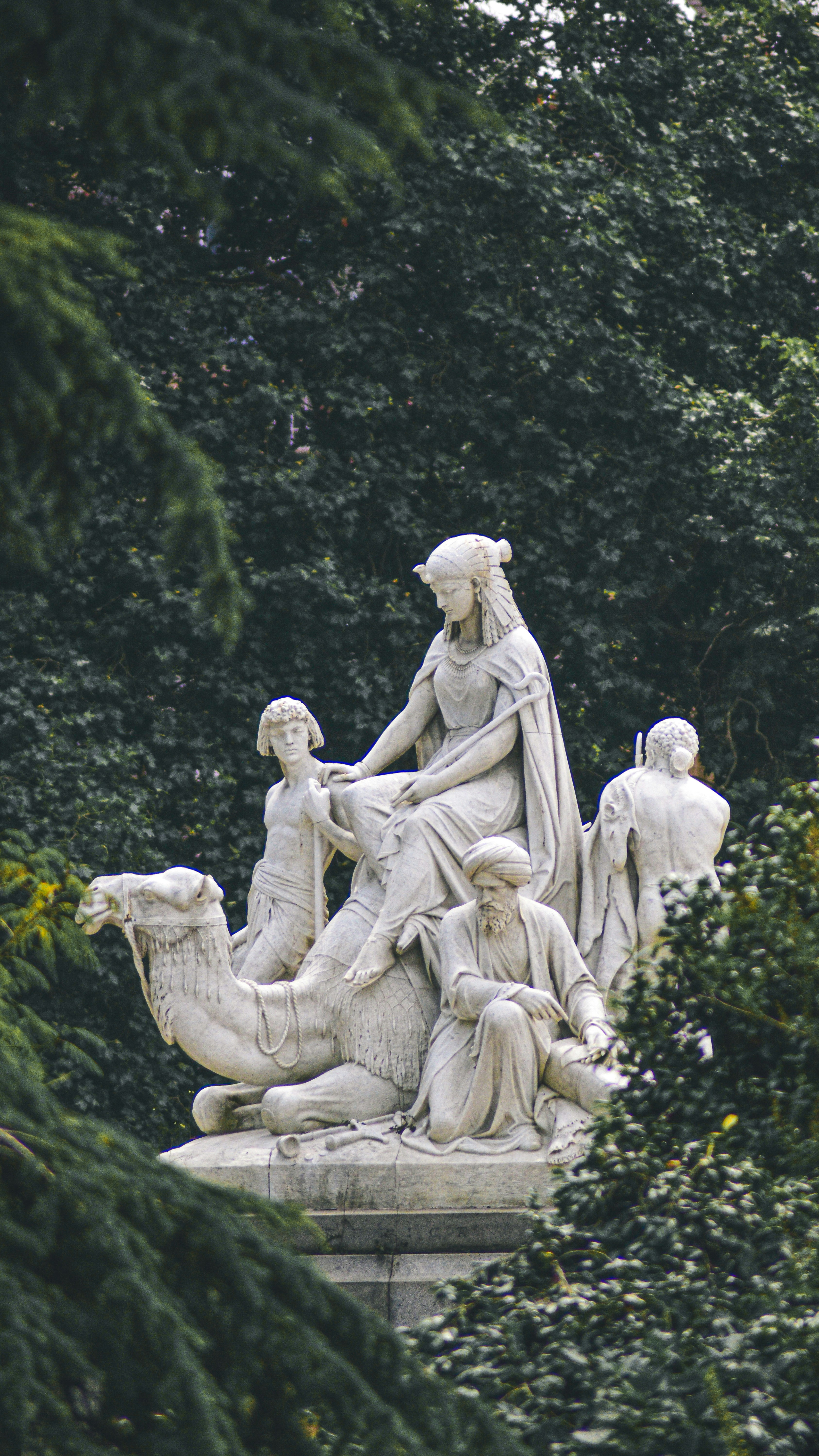 Intricate marble sculpture depicting a woman on a camel accompanied by figures, set against a lush green backdrop.