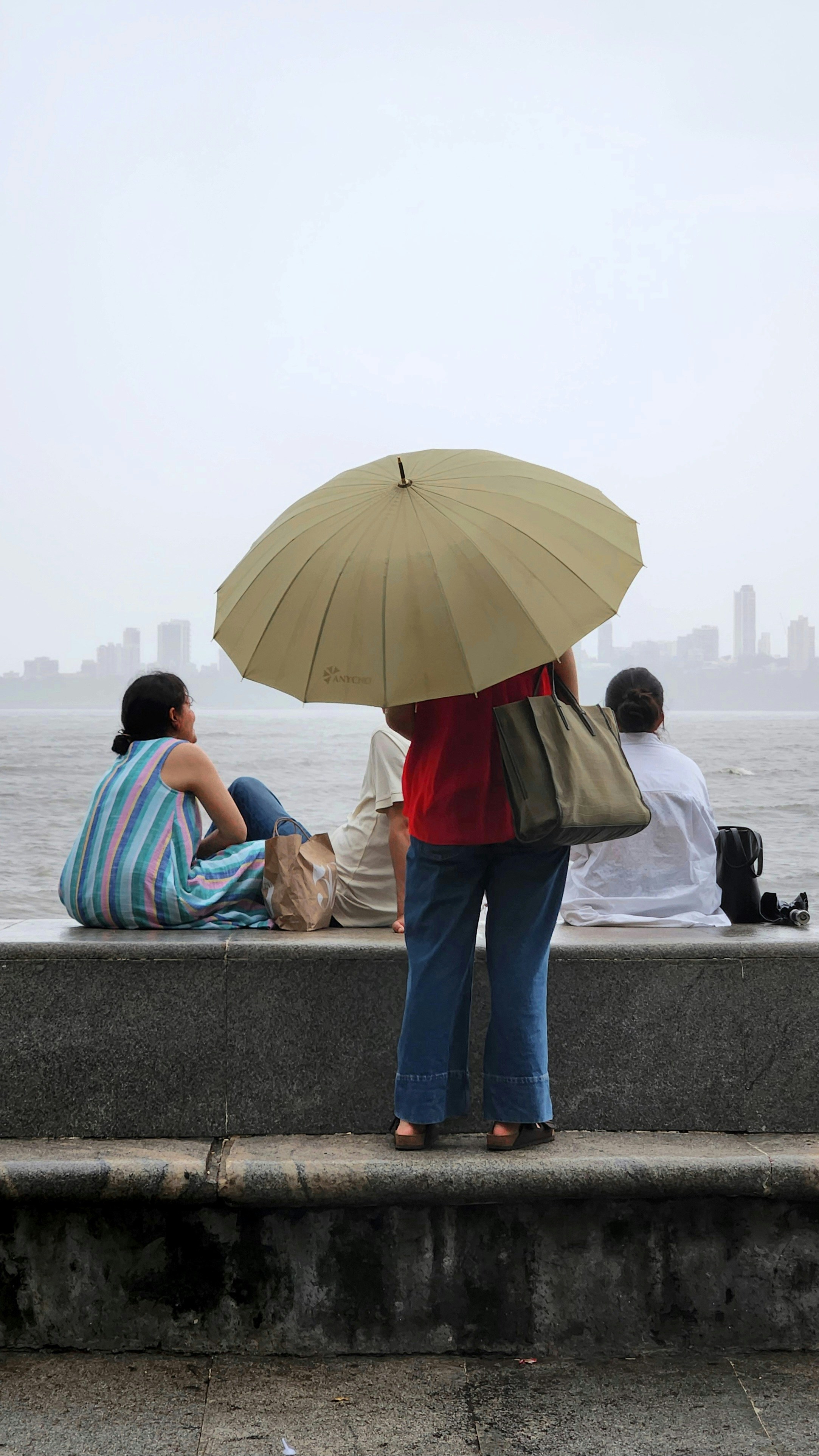 Group of individuals sitting by the waterfront, with one person holding a large umbrella, creating a serene atmosphere amidst the urban skyline.