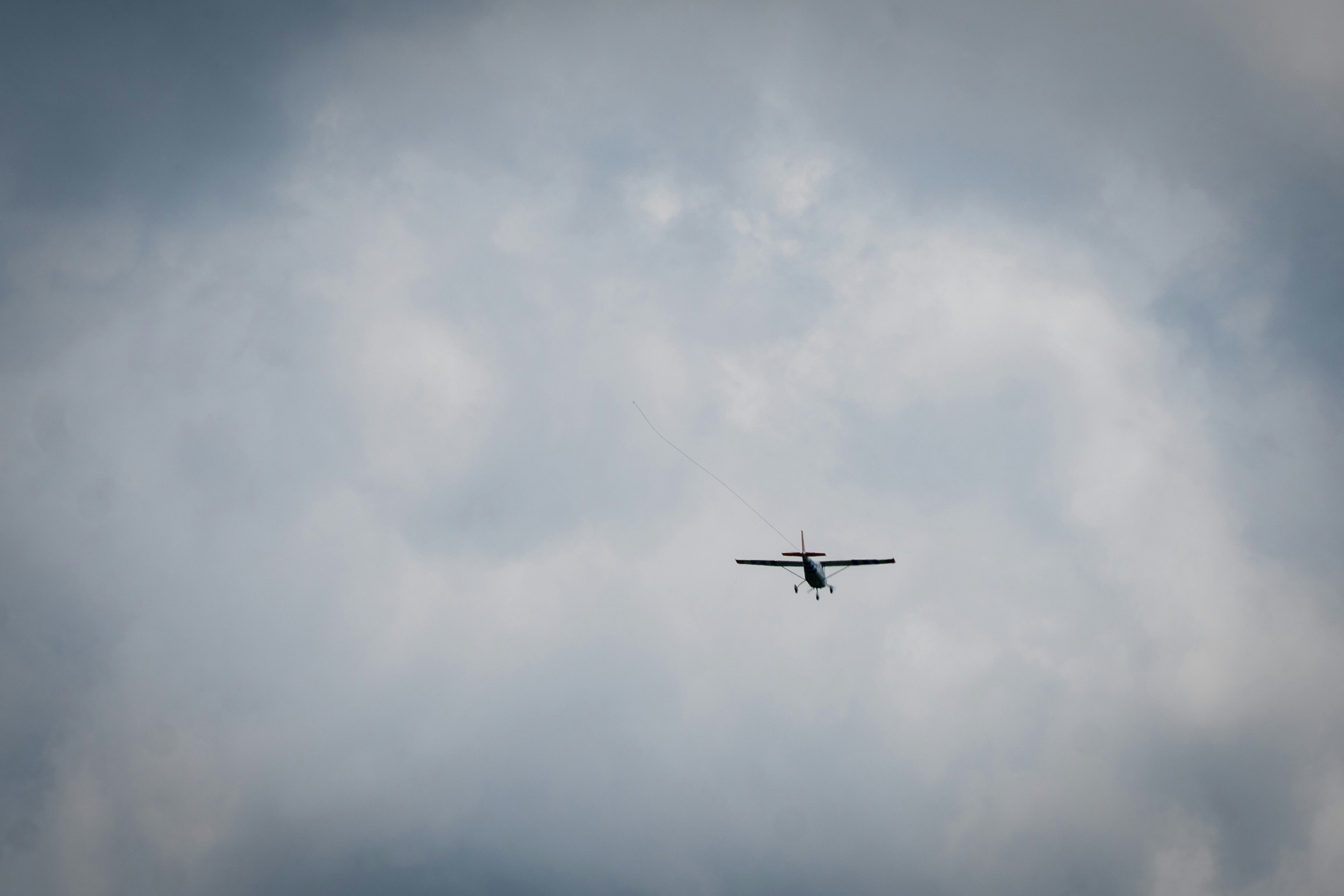 A small tow plane ascends into a moody sky, trailing a long cable through thick, textured clouds. The image captures a quiet tension between the mechanical and natural, as the aircraft carves a path through the stormy atmosphere. | Airplane flies through the cloudy sky.