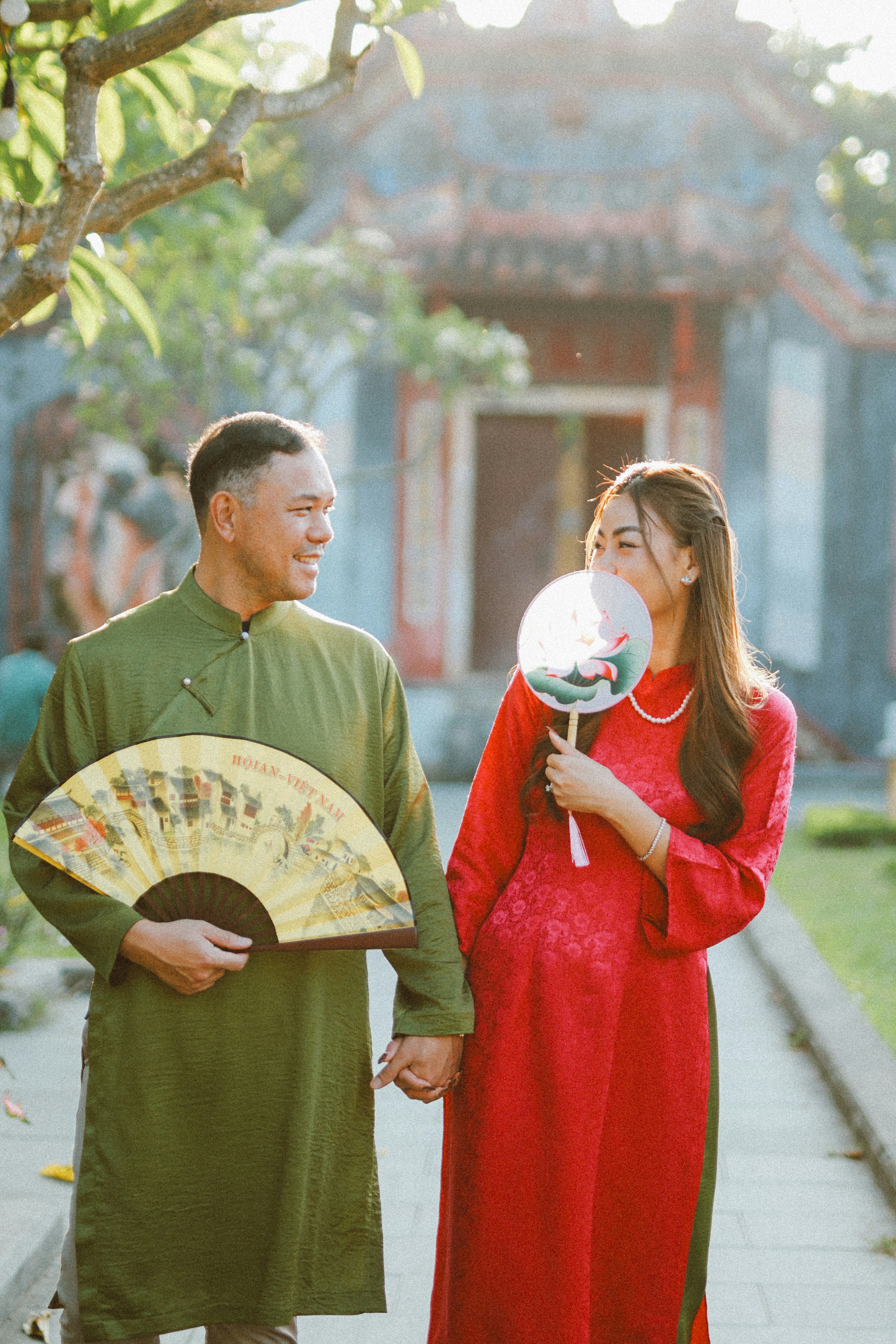 Couple in traditional attire holding hands, surrounded by a serene garden setting. The man holds a decorative fan while the woman playfully holds a lantern.