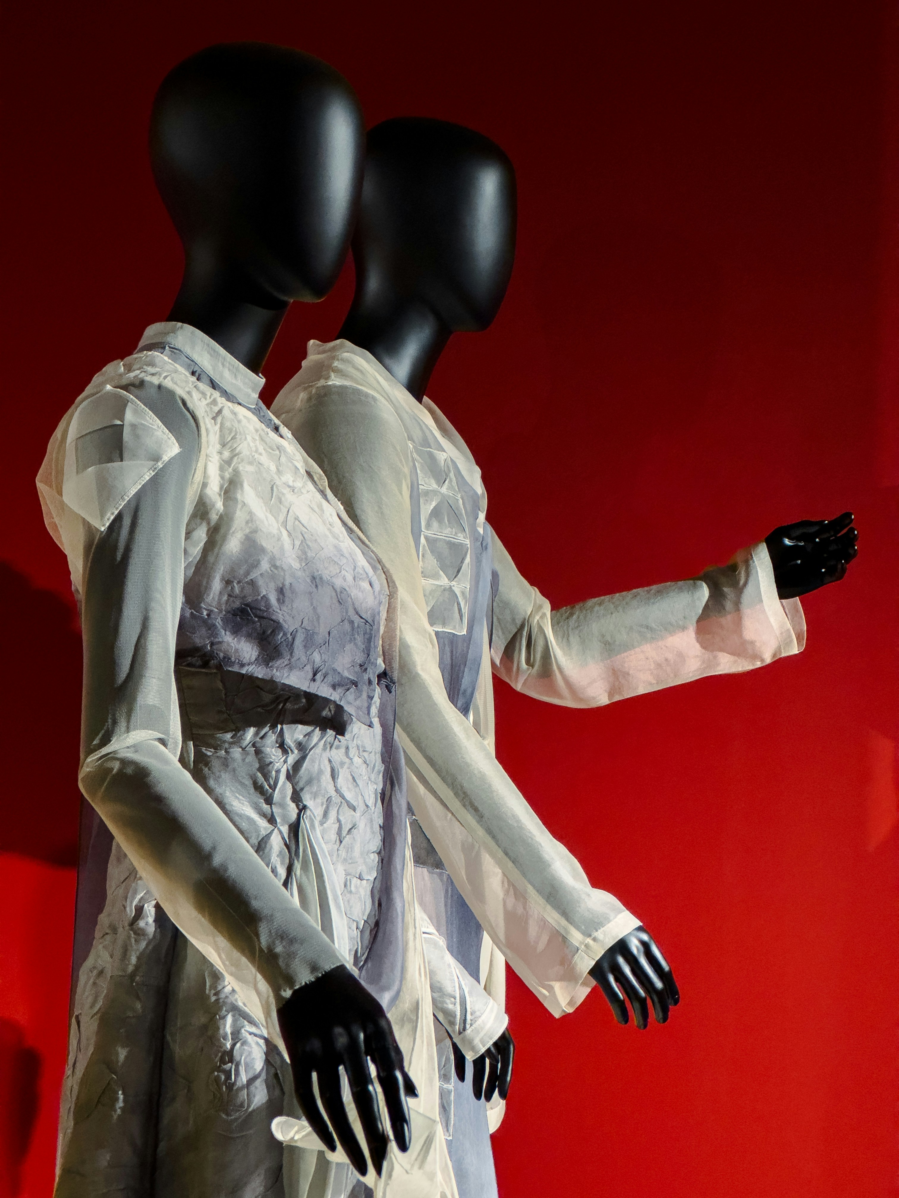 Two mannequins dressed in translucent garments against a vibrant red backdrop, showcasing modern fashion design elements.