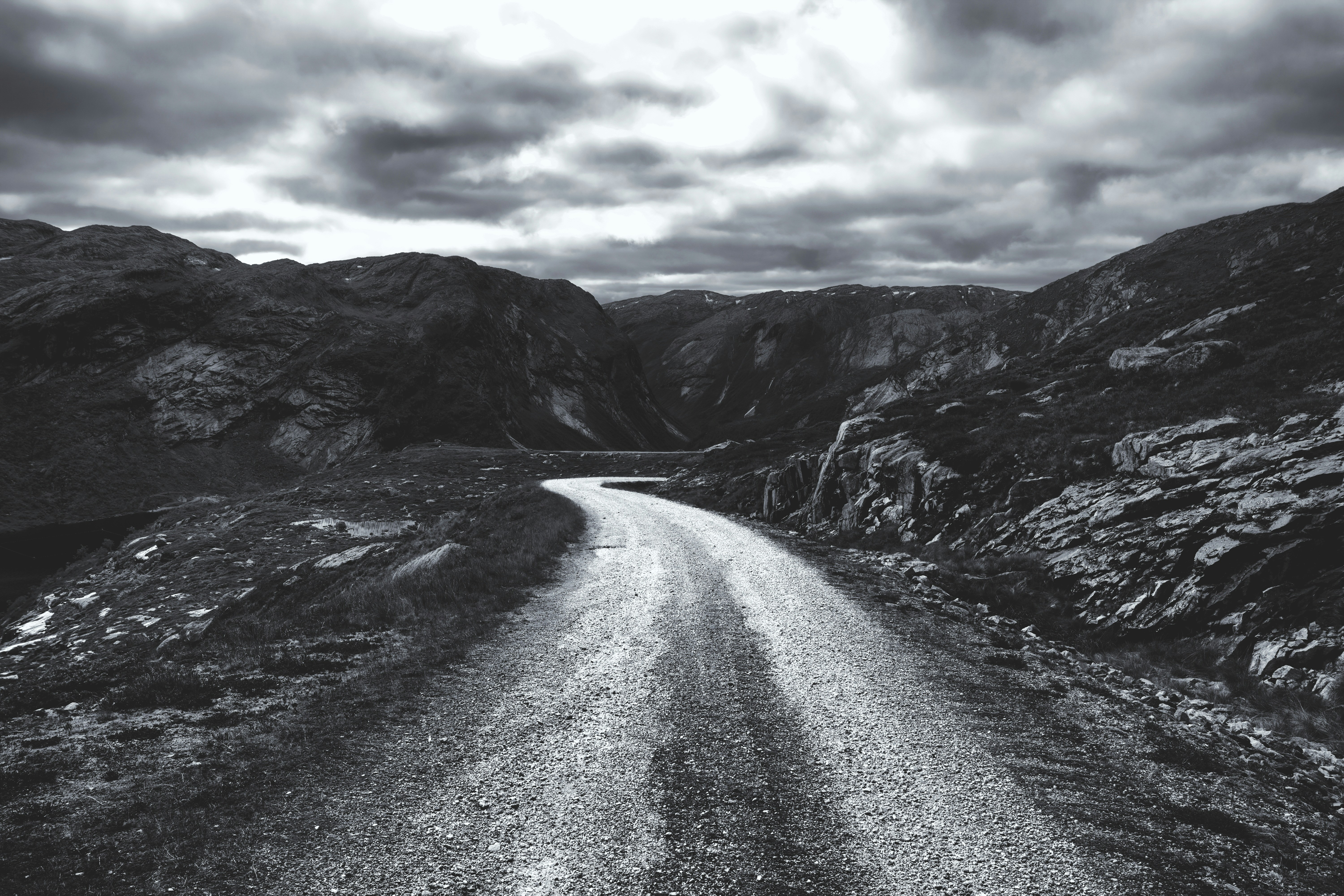 A gravel road meanders through a dramatic mountainous terrain under a brooding sky. The stark contrasts highlight the rugged beauty of the natural surroundings.