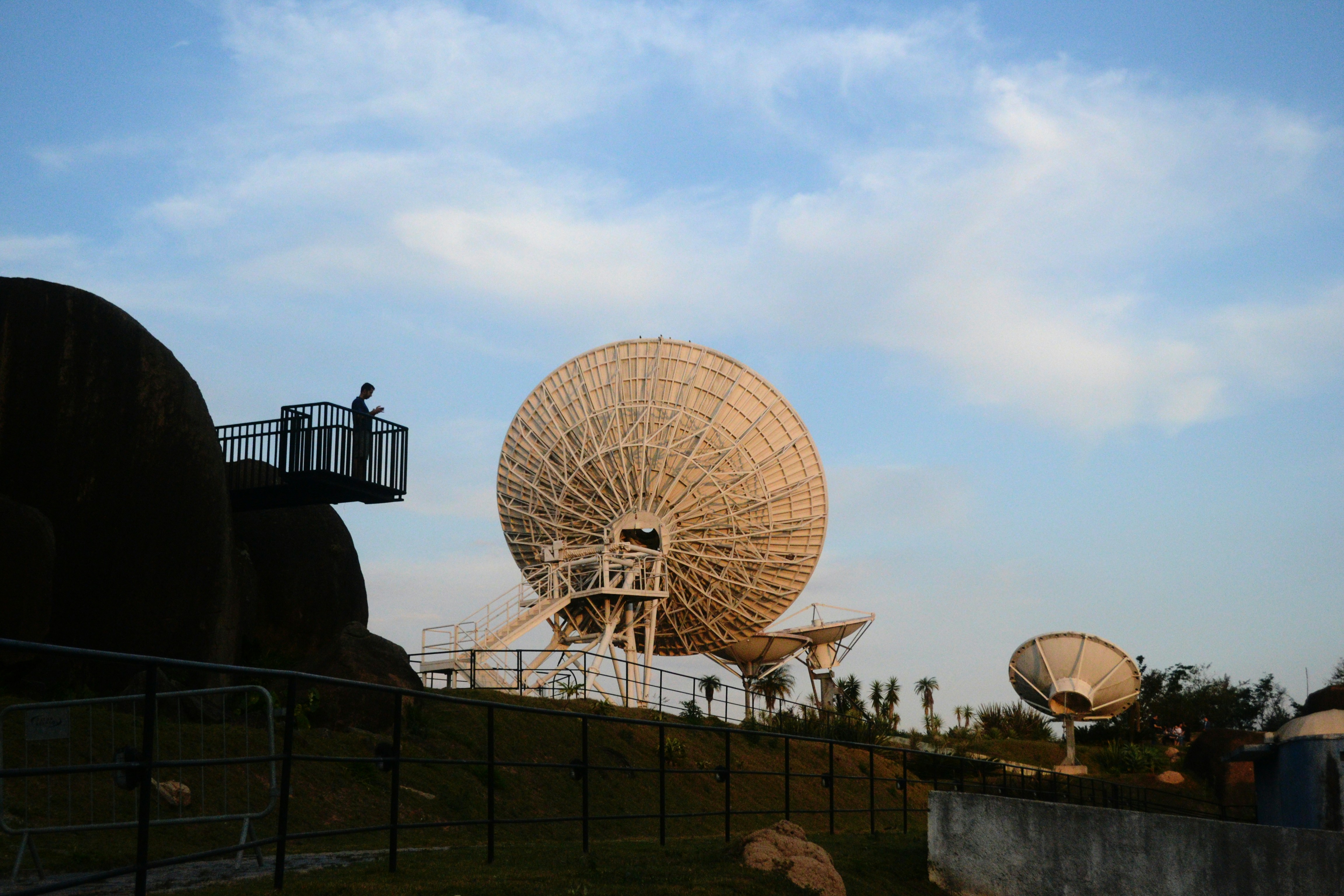 Silhouette of a person on a platform with large satellite dishes in the background, under a blue sky with wispy clouds.