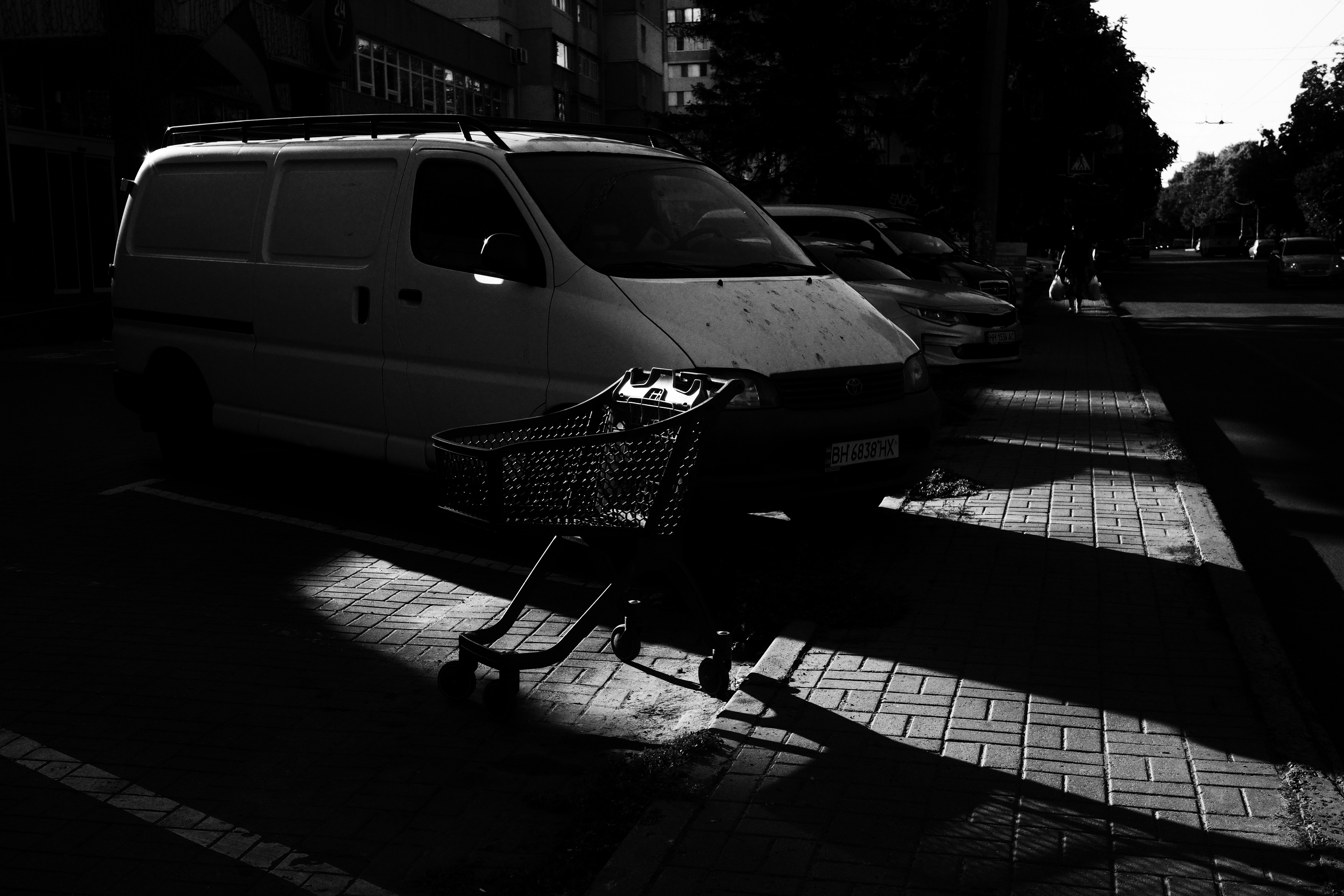A stark black and white image captures a solitary shopping cart in the foreground, casting a long, textured shadow across the patterned sidewalk. Behind it, a white van is parked, partially obscured by shadows, with other vehicles visible further down the street. The scene, likely set in Odesa, is characterized by dramatic contrasts between light and deep shadow, emphasizing the urban environment and the seemingly abandoned cart. | A van and shopping cart sit in shadow.