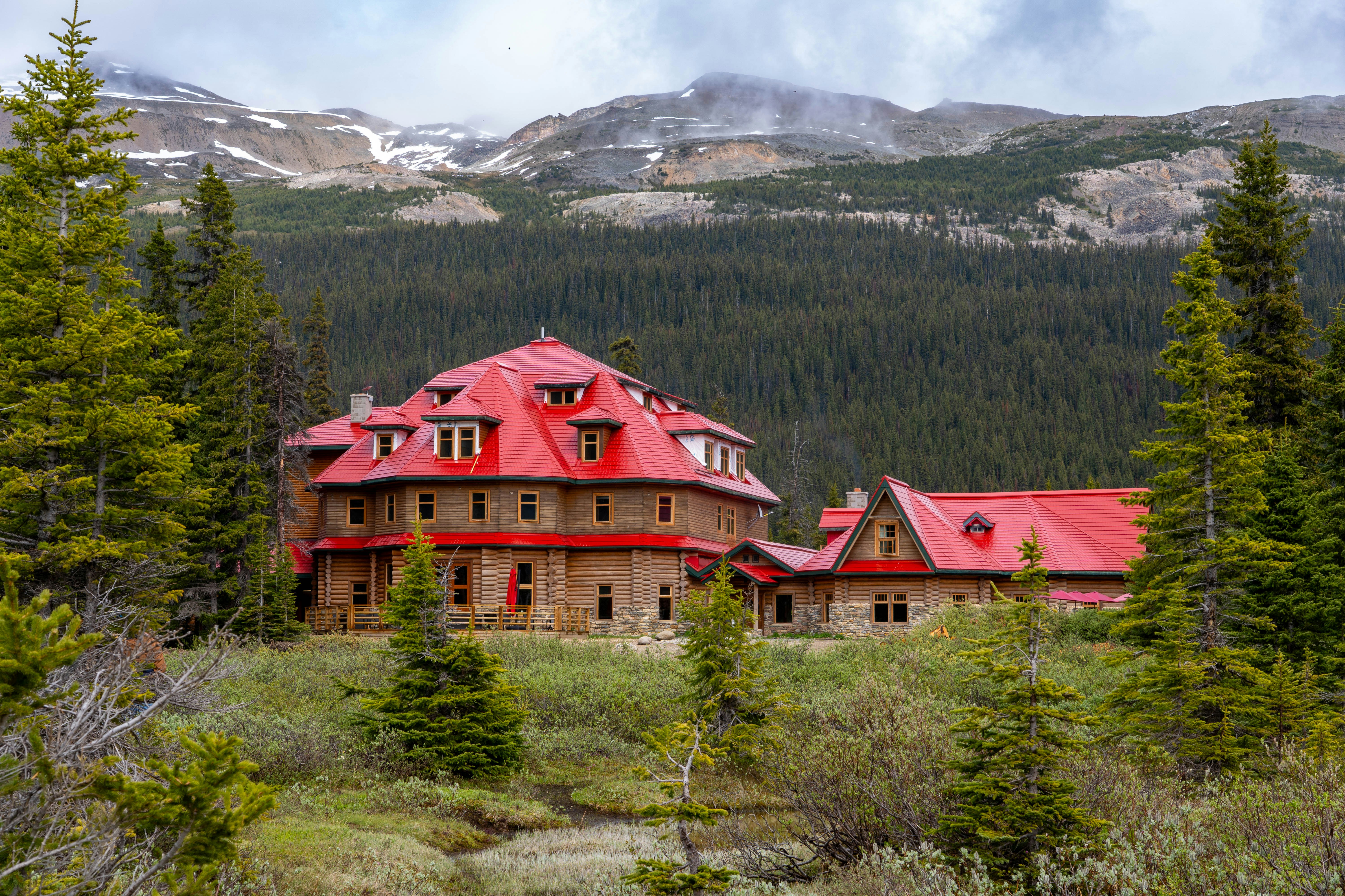 Historic lodge with striking red roofs nestled in a lush green landscape, framed by towering mountains and a dramatic sky.
