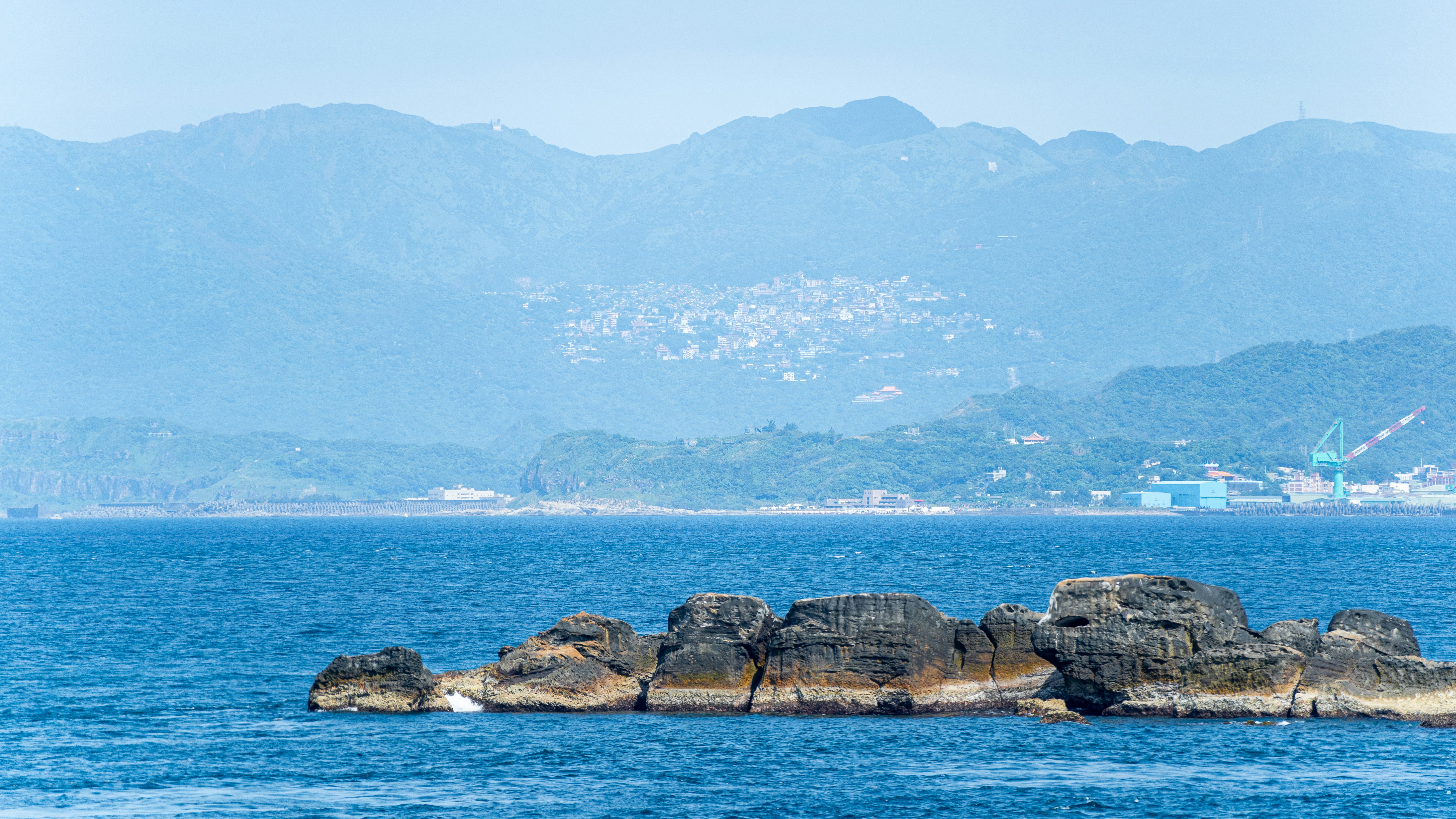Rocky island sits in the ocean, mountains in the distance.