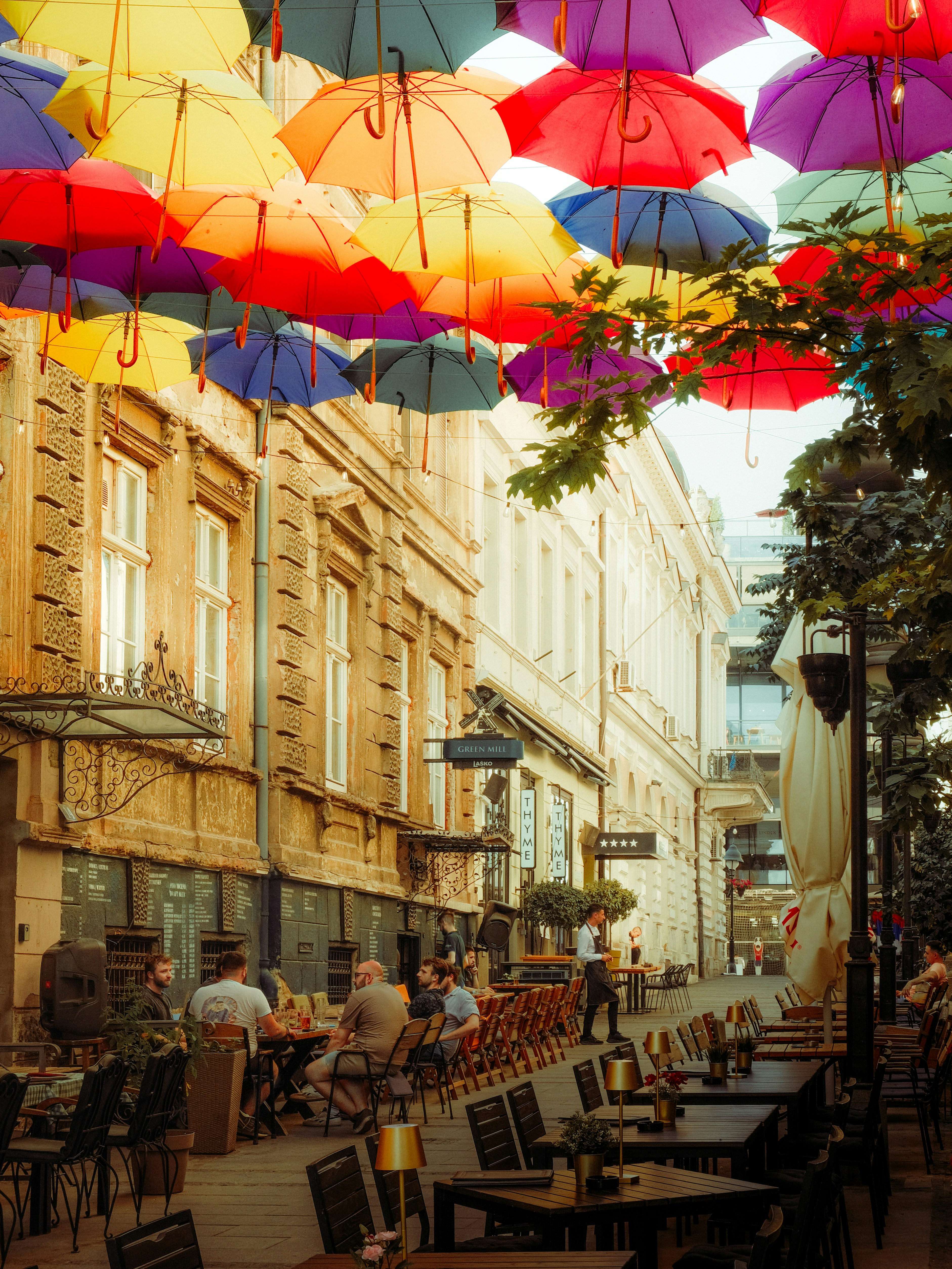 Colorful umbrellas suspended above a quaint street lined with outdoor cafes and historic buildings. The scene captures a lively atmosphere in an urban setting.