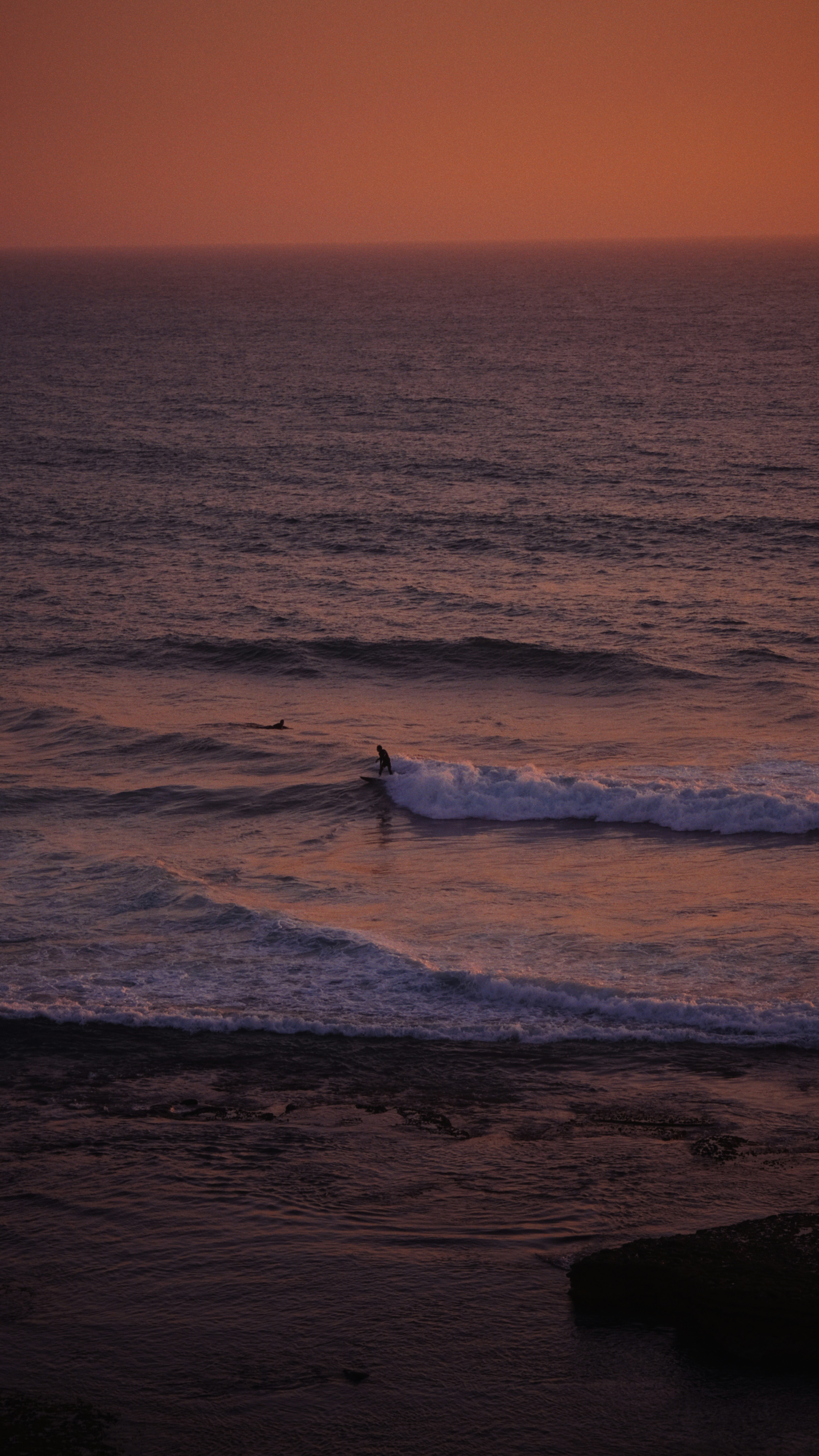 Surfer navigating gentle waves under a gradient sky at dusk, with another surfer visible in the distance. 