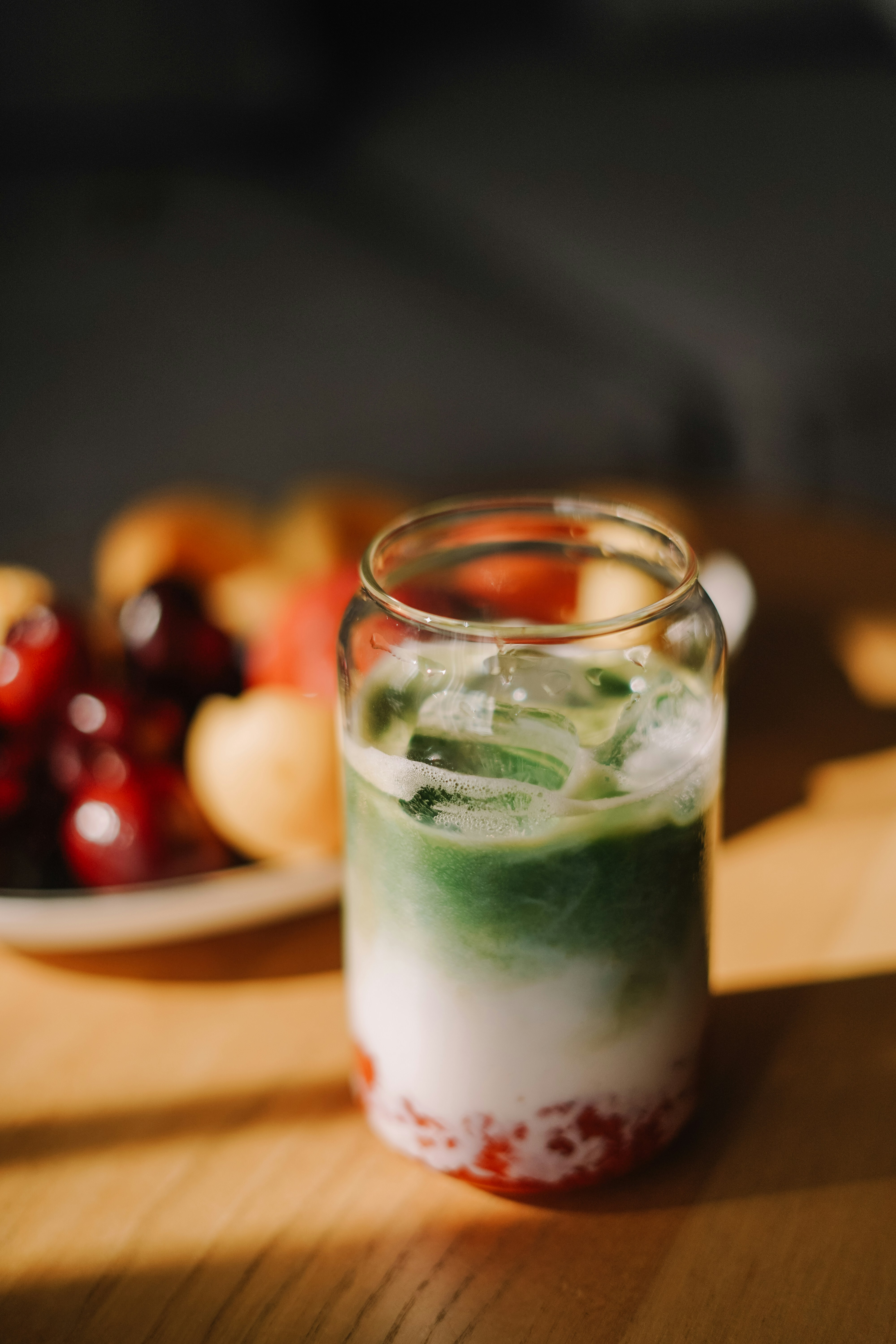 Vibrant drink with layers of green, white, and red served in a clear glass, accompanied by a plate of assorted fruits. The sunlight highlights the textures and colors beautifully.