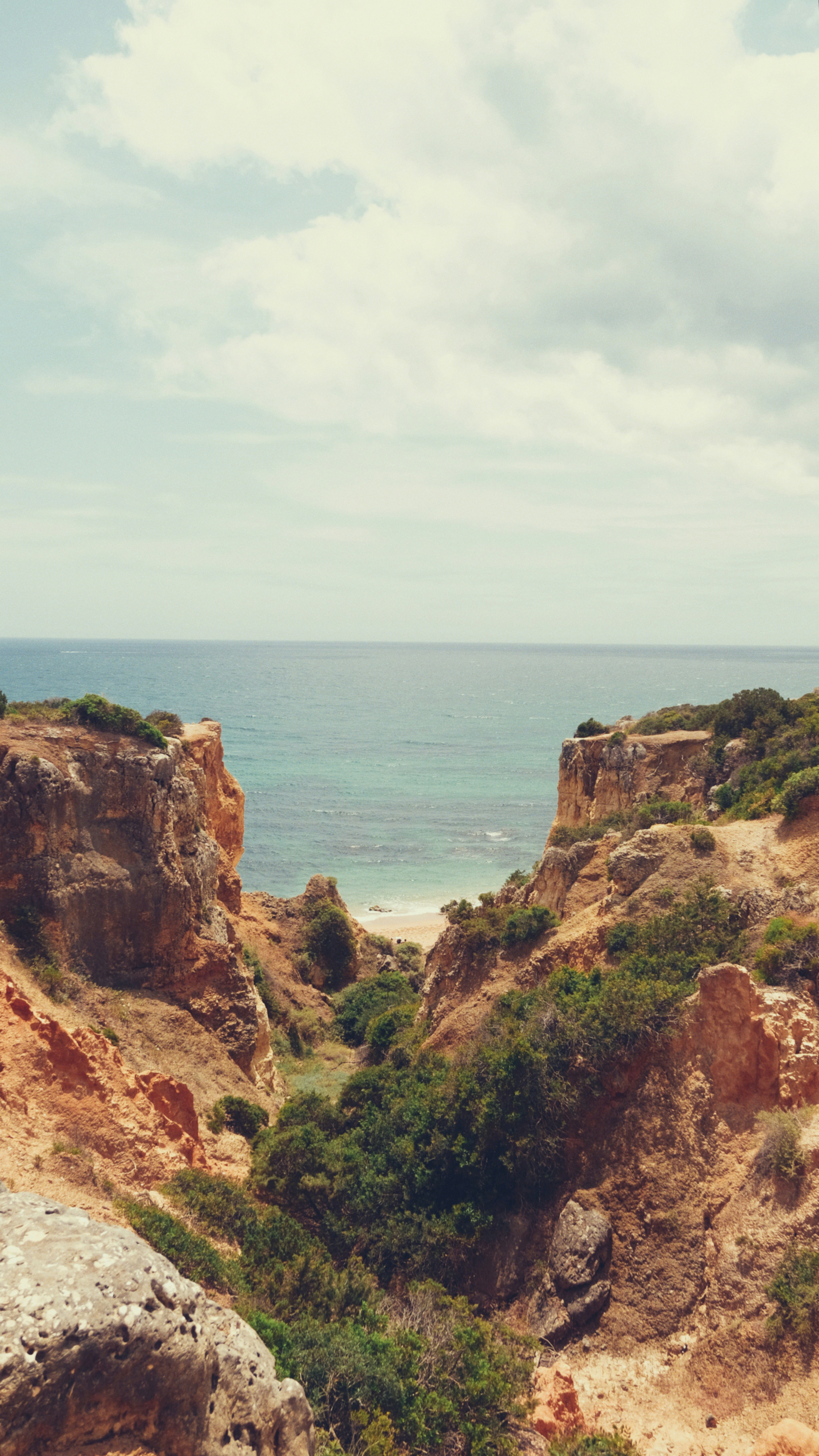 A breathtaking view of a coastal landscape with rugged cliffs framing a tranquil ocean scene. Lush greenery contrasts with the sandy shore below.
