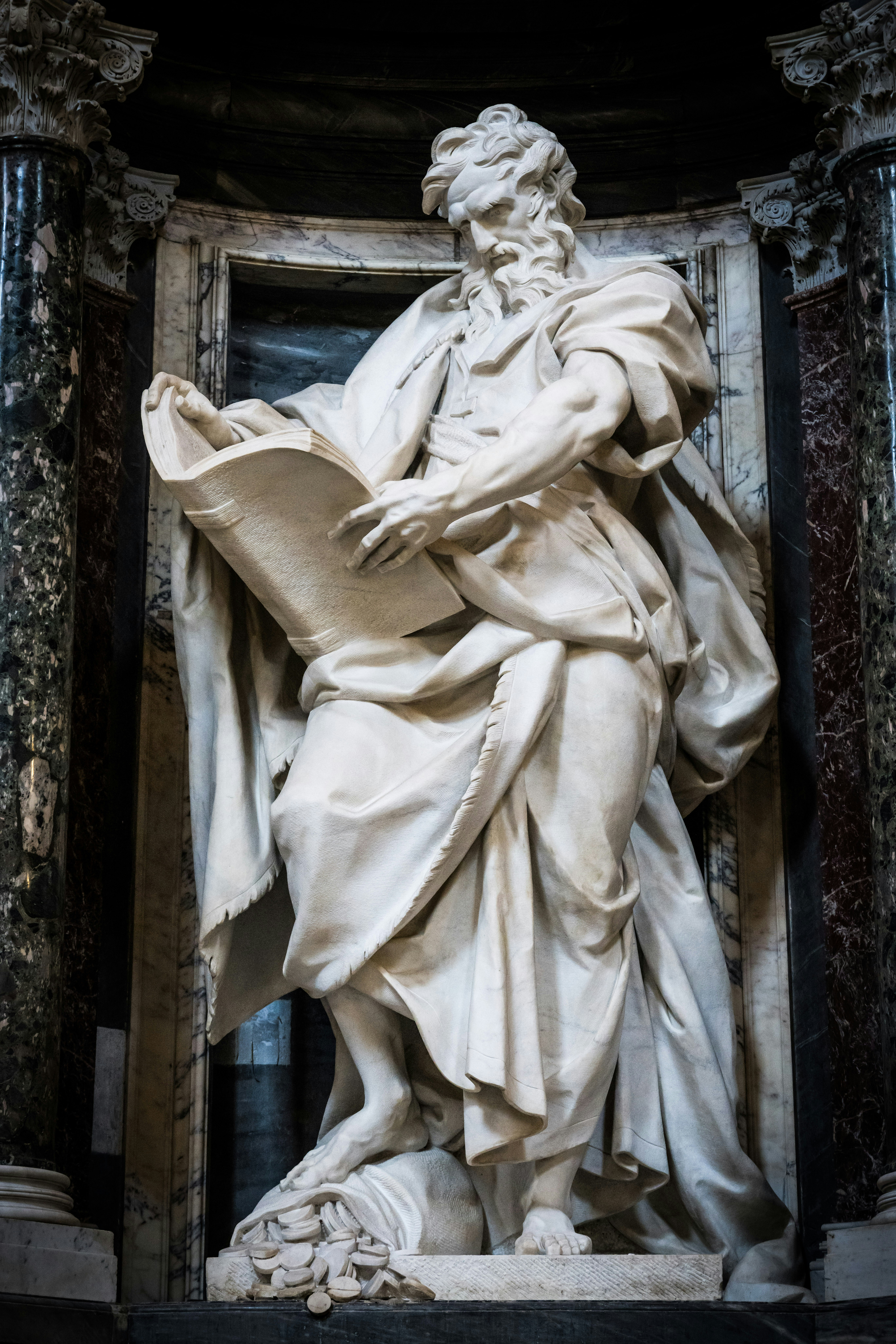 Marble statue of a figure holding an open book, draped in flowing robes, set against a richly decorated backdrop.