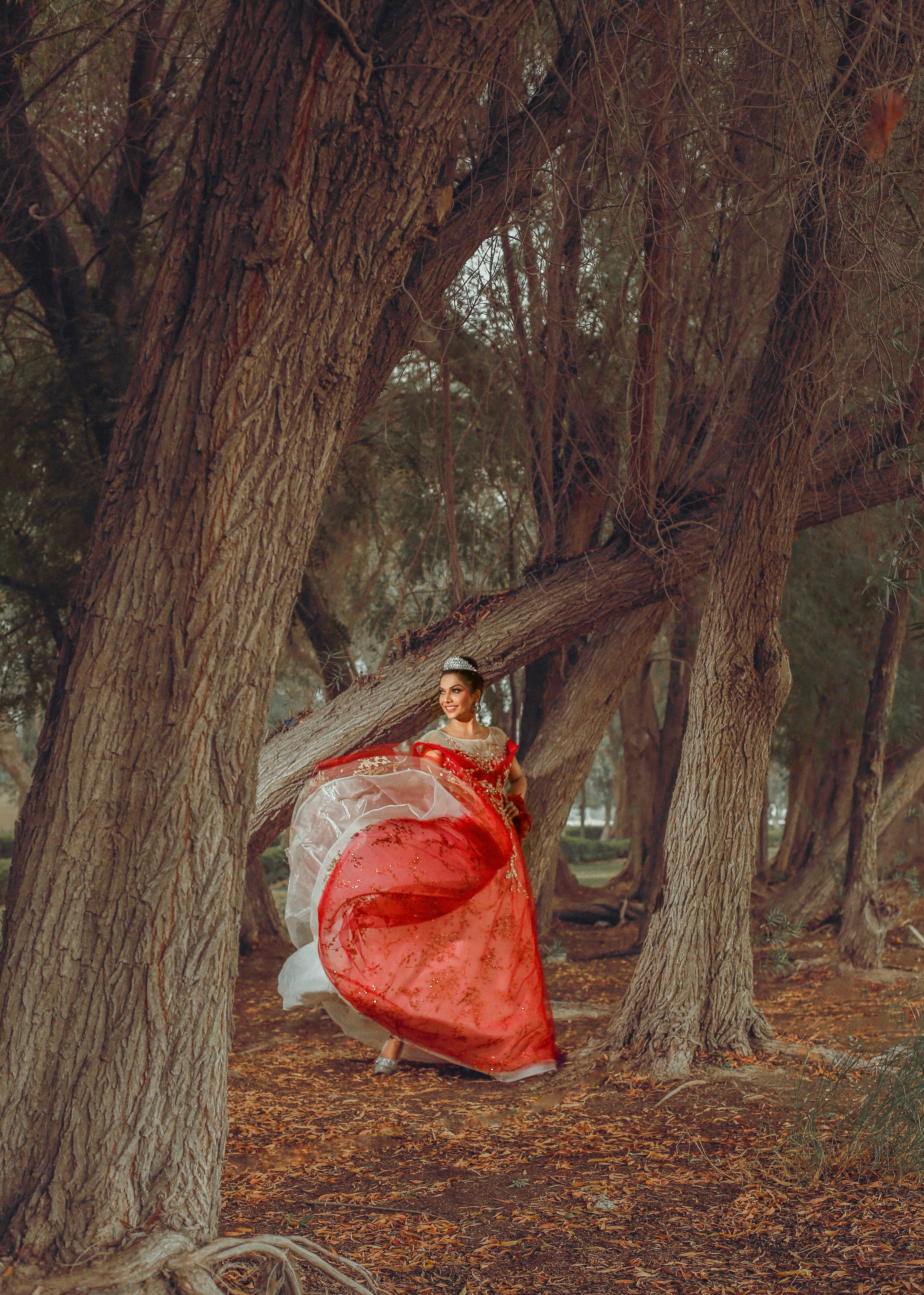 A captivating fashion portrait captured by Naeem Ad, featuring a young woman in a stunning red gown posing gracefully in a dense, dreamy forest. The rich textures of nature combined with the elegance of the dress create a cinematic and storybook aesthetic. Shot with attention to color, light, and composition — perfect for editorial, fashion, or fairy-tale themes. 📸 Photo by Naeem Ad Studios | @naeem_ad_studios1 | Woman in red dress poses amidst trees.