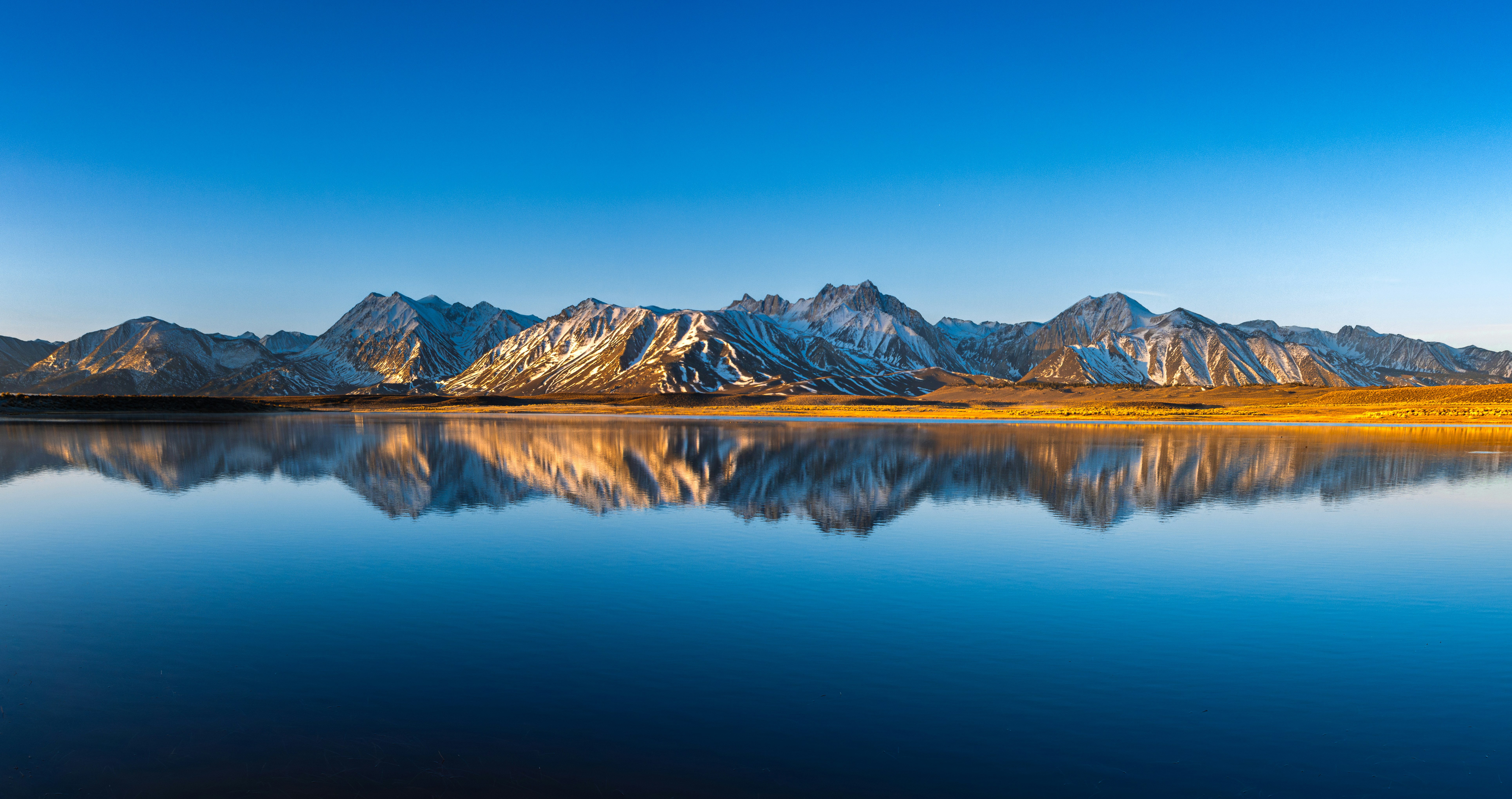 Snow-capped mountains reflecting in a tranquil lake under a clear blue sky.