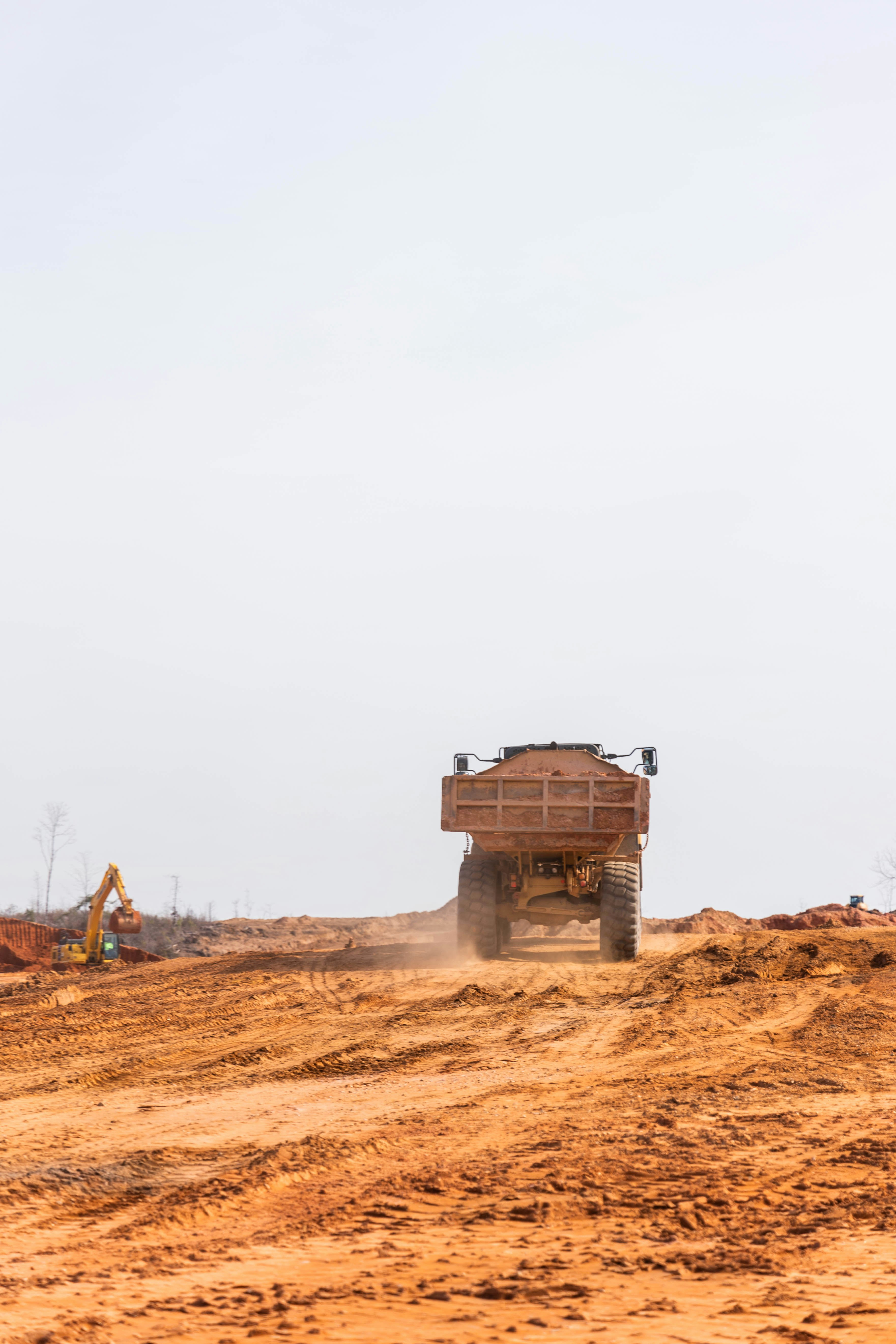 A large dump truck navigates a dusty construction site, leaving trails in the orange earth. An excavator is visible in the background, indicating ongoing excavation work.