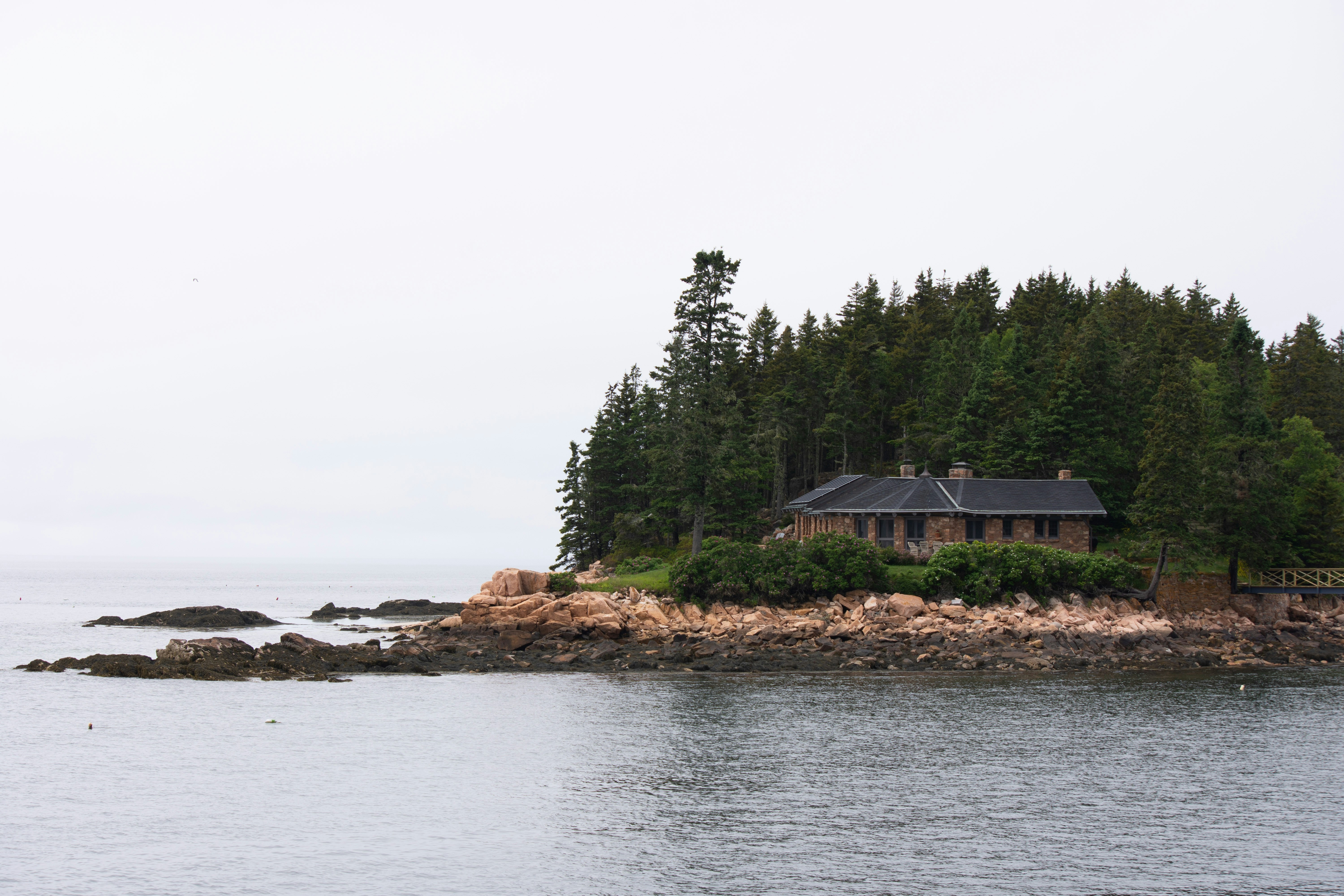 A charming stone house nestled on a rocky shoreline, surrounded by lush evergreens and calm waters under a cloudy sky.
