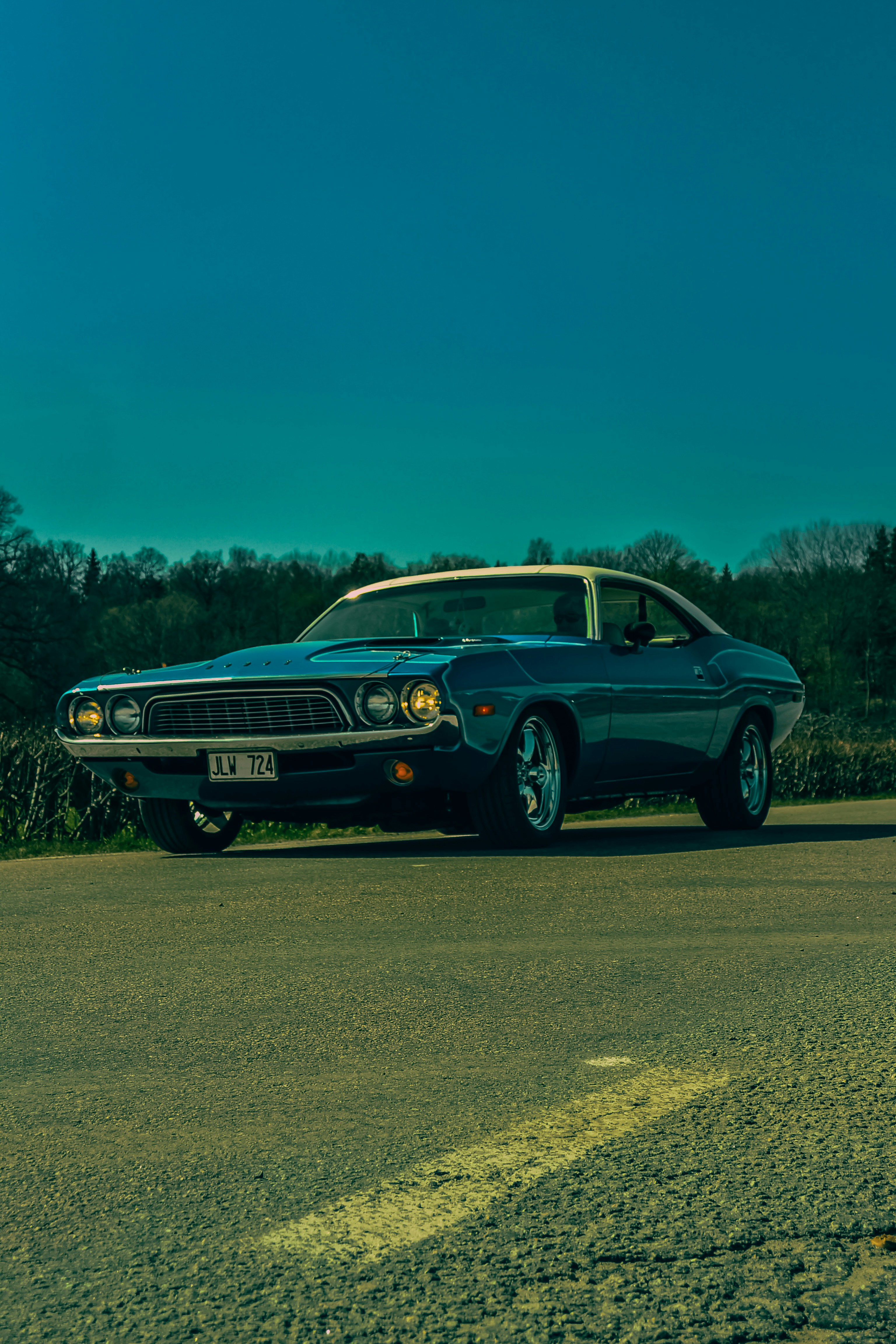 Vintage blue muscle car parked on a sunlit road surrounded by greenery.