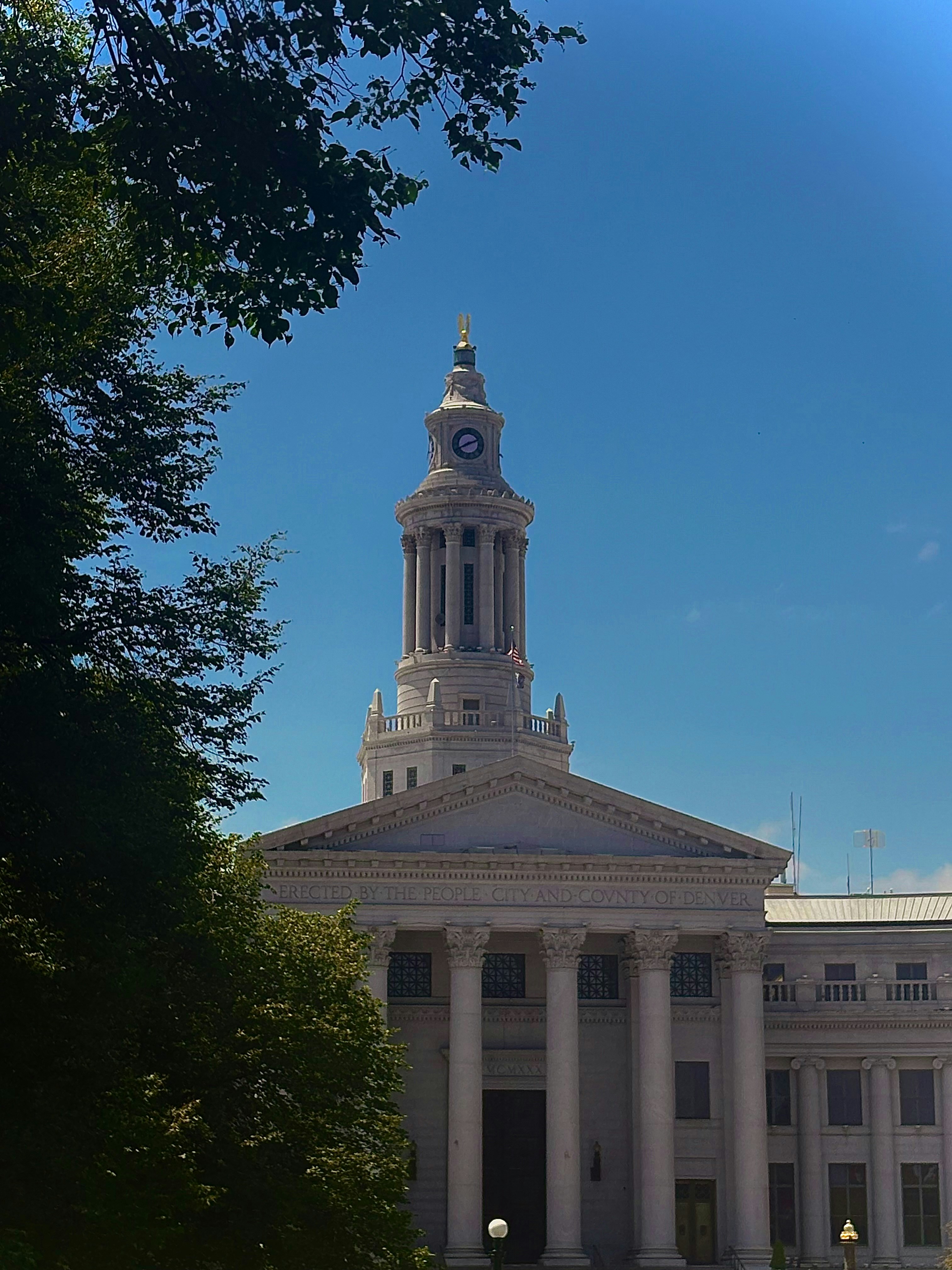 Historic government building with a prominent clock tower, framed by lush greenery under a clear blue sky.