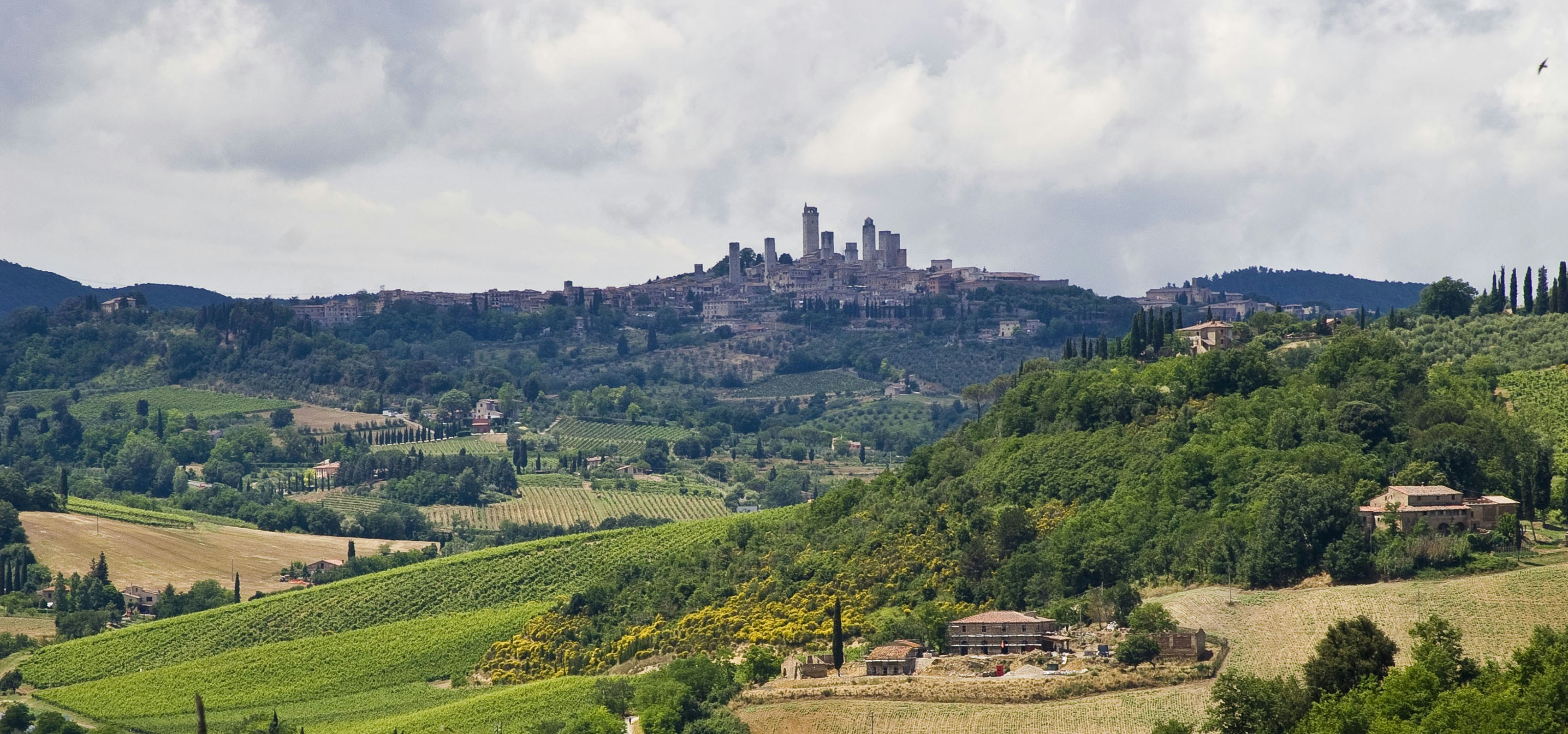 Medieval towers of San Gimignano rise above lush green vineyards and rolling hills under a partly cloudy sky.