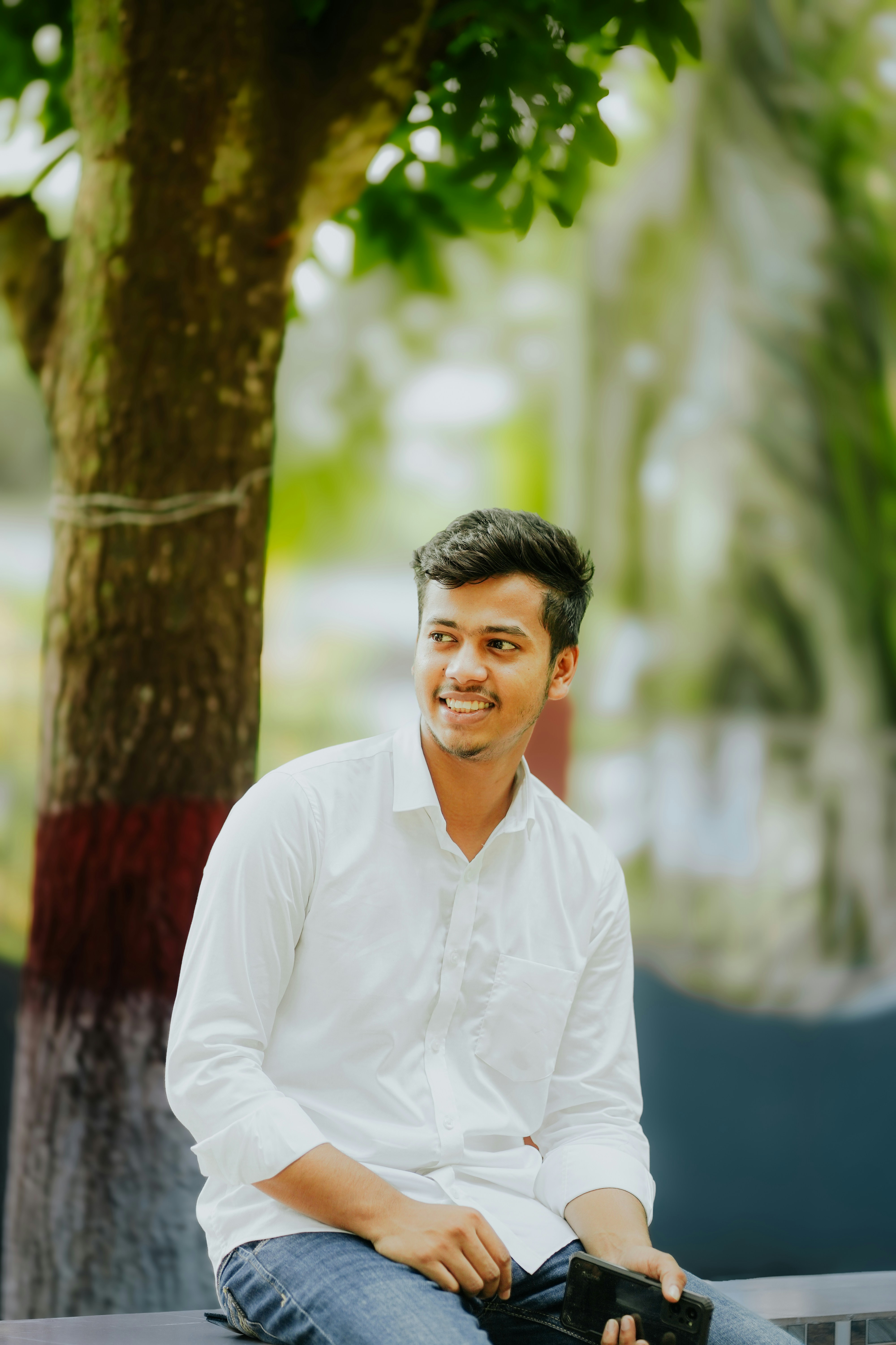 Young man in a white shirt smiling while seated under a tree, holding a phone. Soft natural light filters through the leaves.
