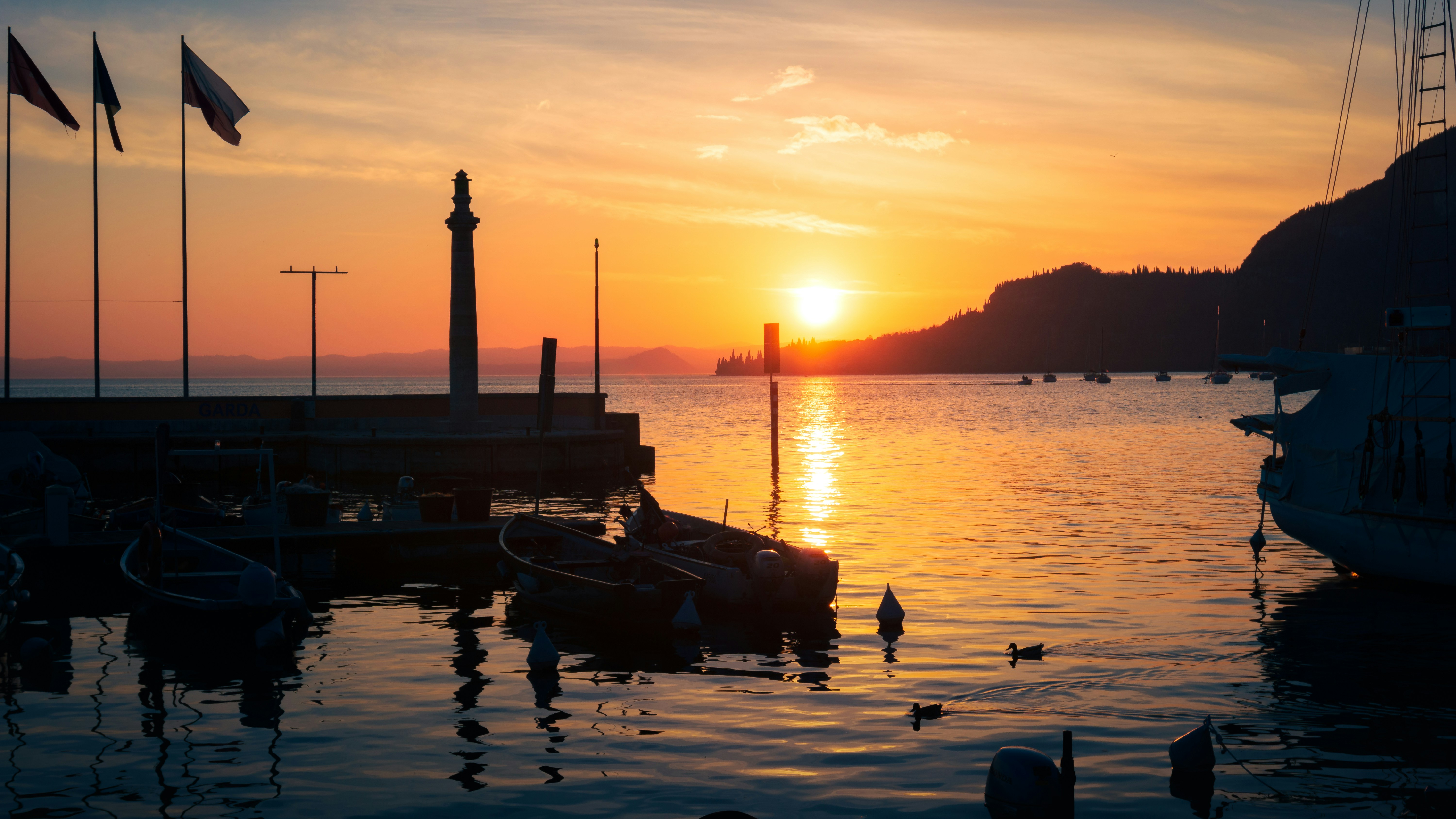 Sunset over a tranquil harbor, with silhouettes of boats and flags against a vibrant sky. The warm hues reflect beautifully on the water's surface.