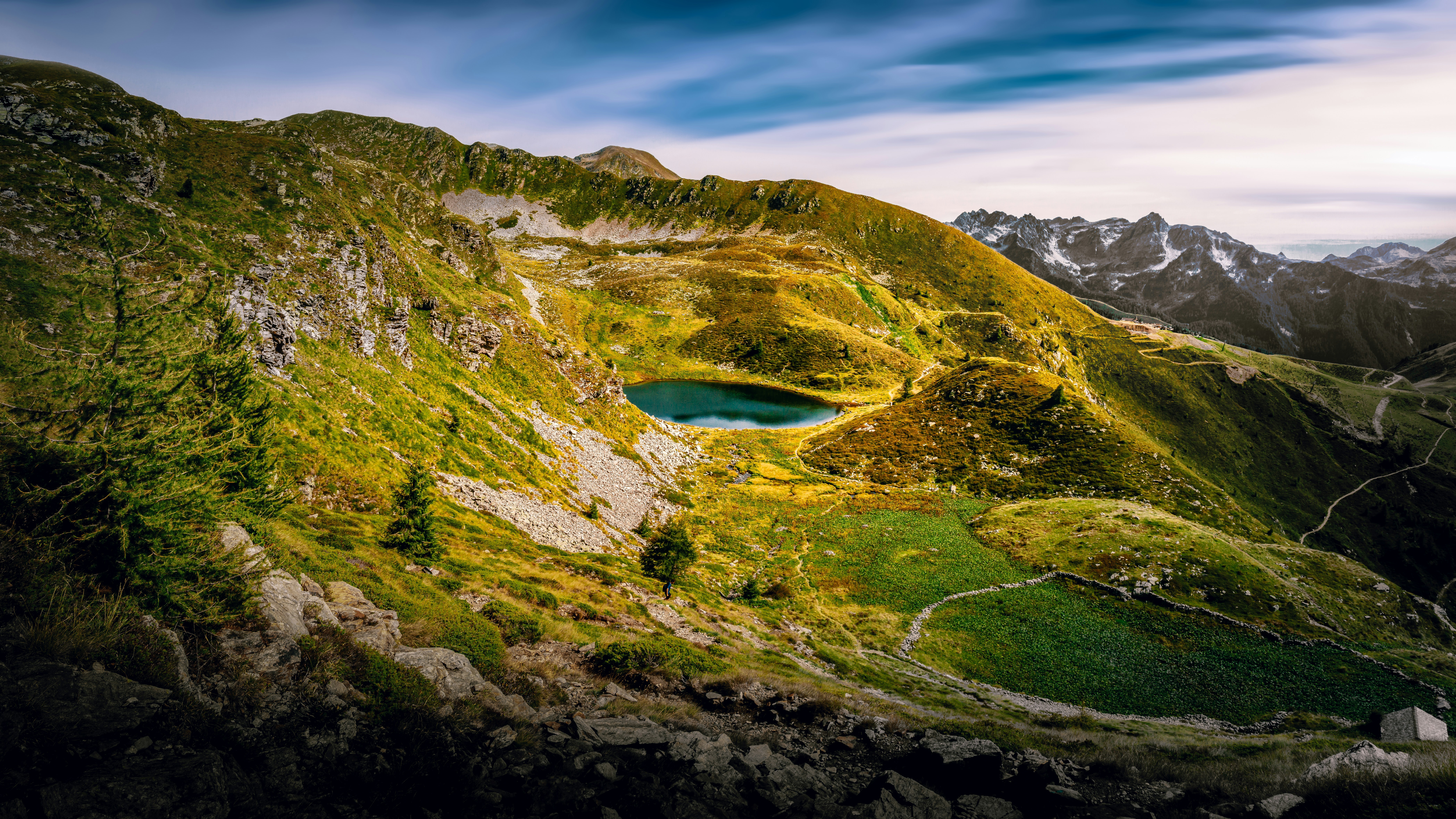 Vibrant green landscape surrounding a tranquil blue lake, nestled within a mountainous terrain under a dynamic sky.