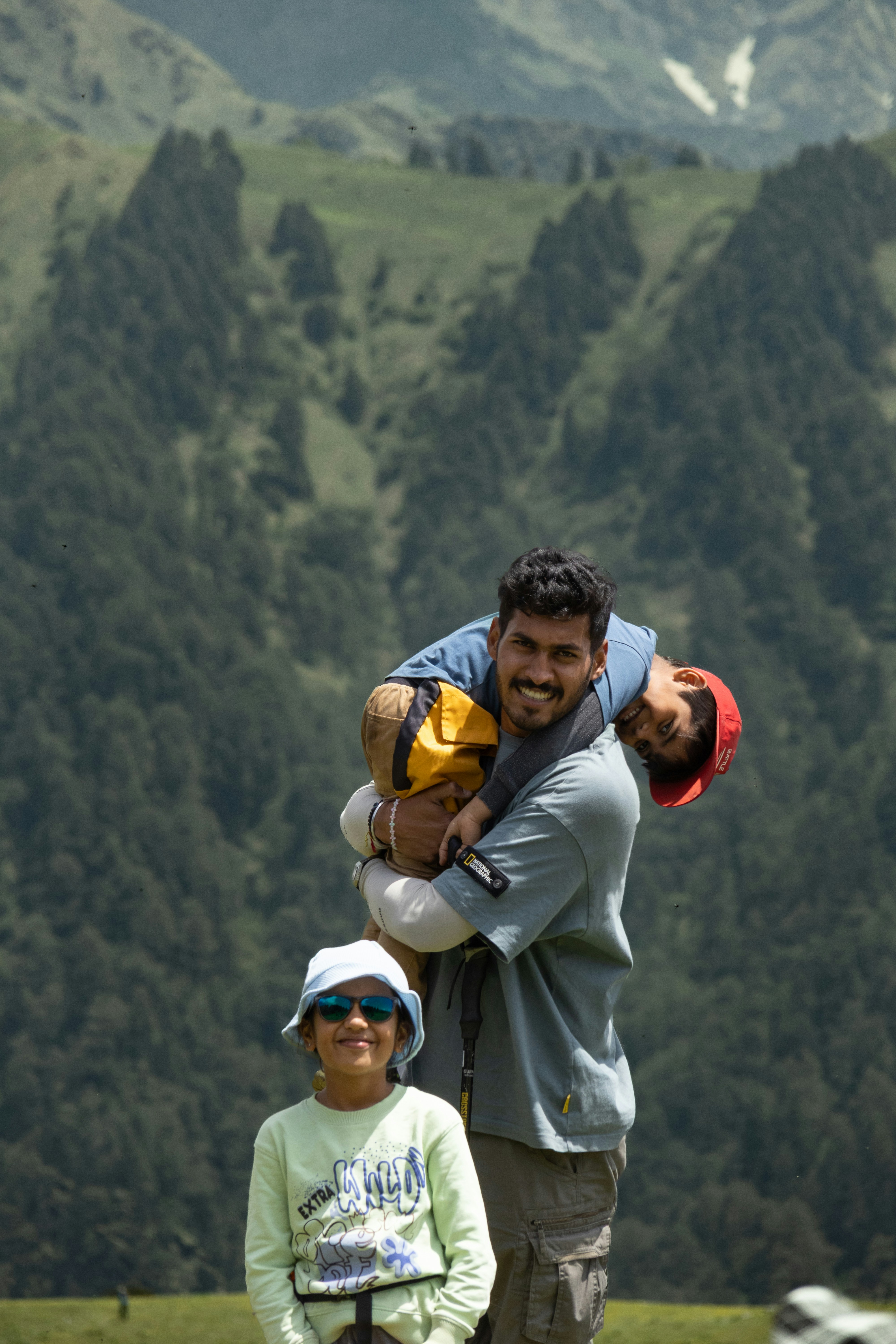 A man joyfully carries a child on his back while a girl stands in front, all set against a lush green mountainous backdrop.